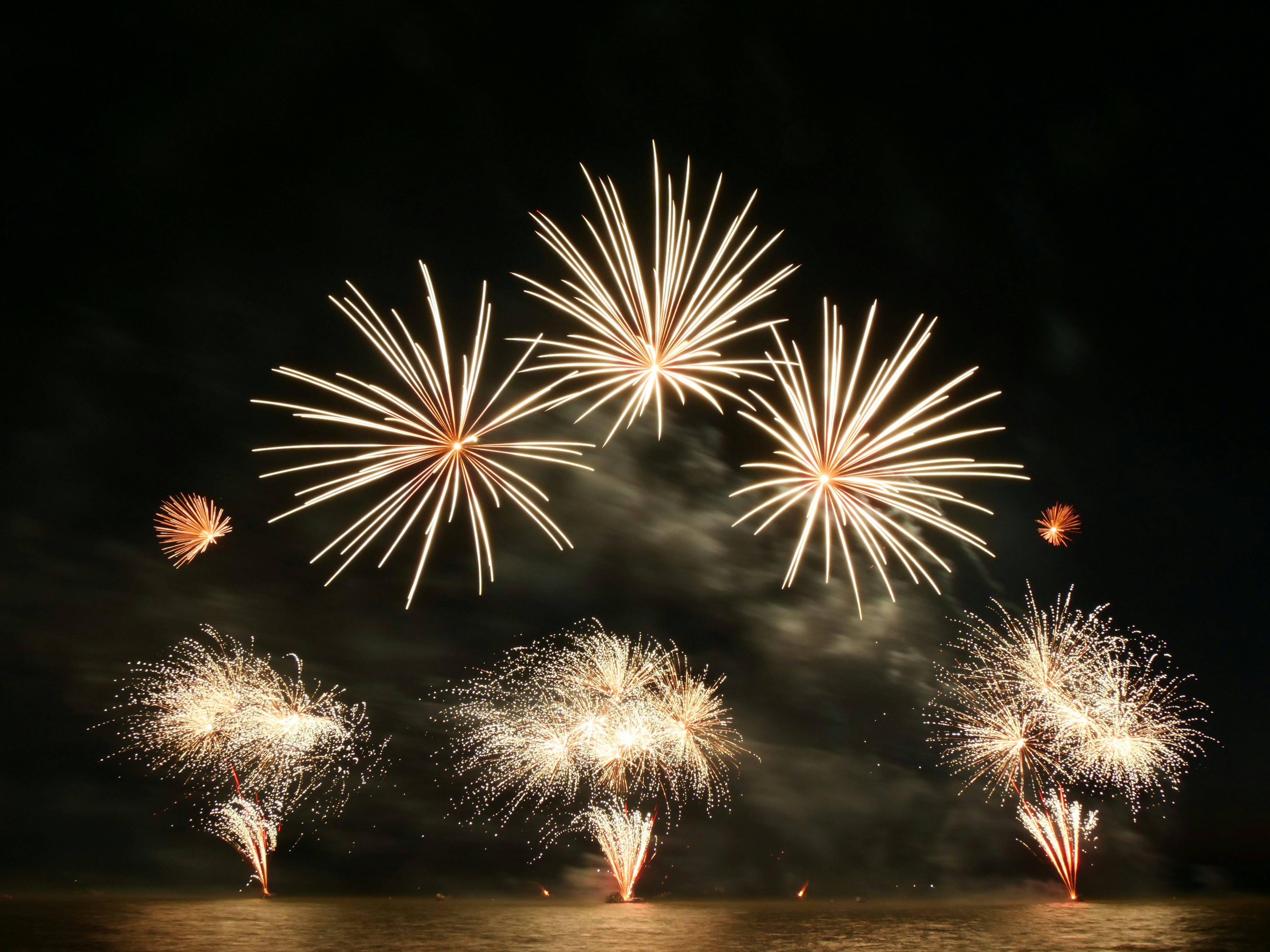 Five golden fireworks bloom across a dark sky above a quiet shoreline, with trailing sparks and smoke forming a layered display.