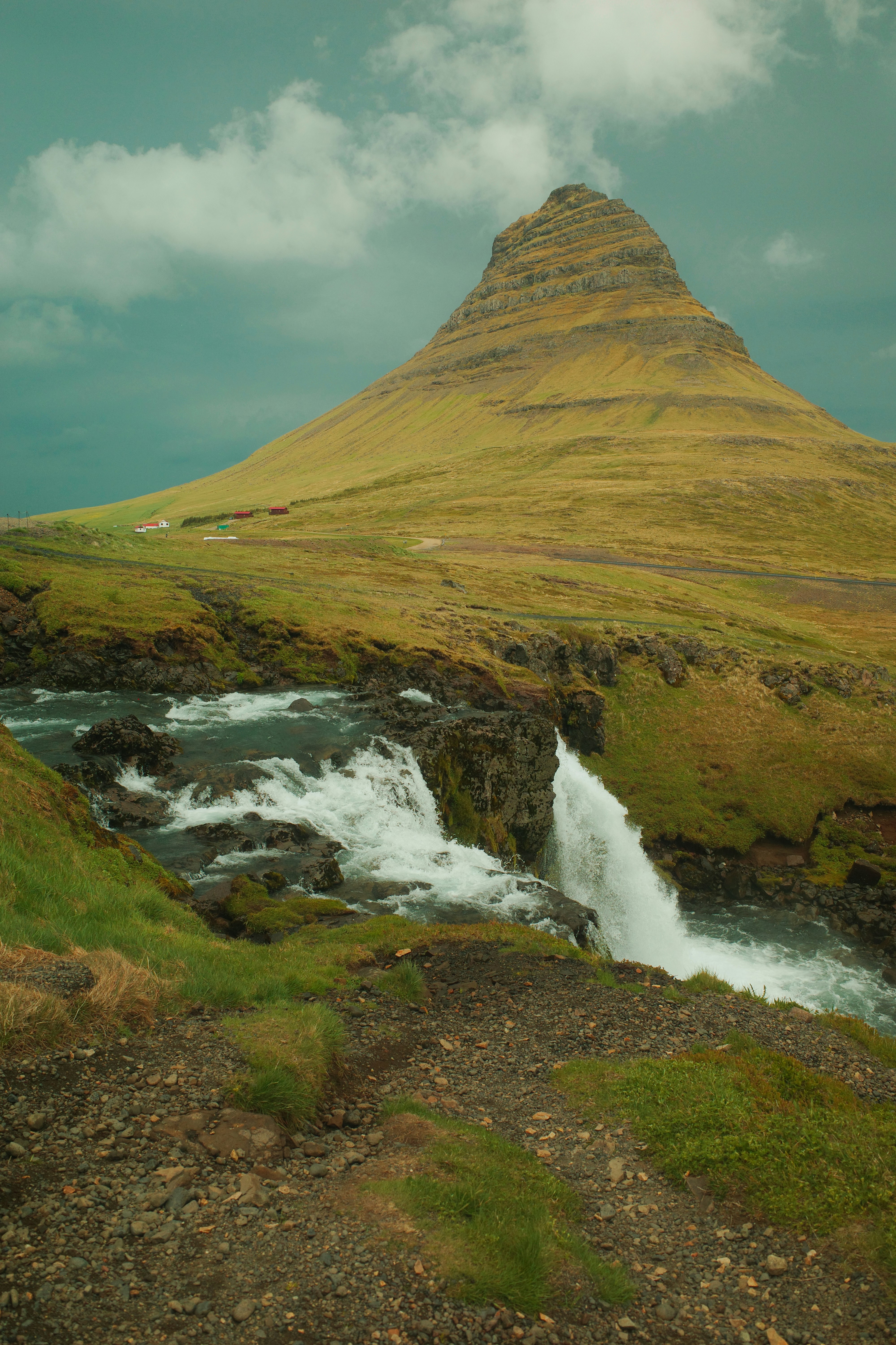 A lone cone of rock rises over emerald meadows and a rushing shoreline