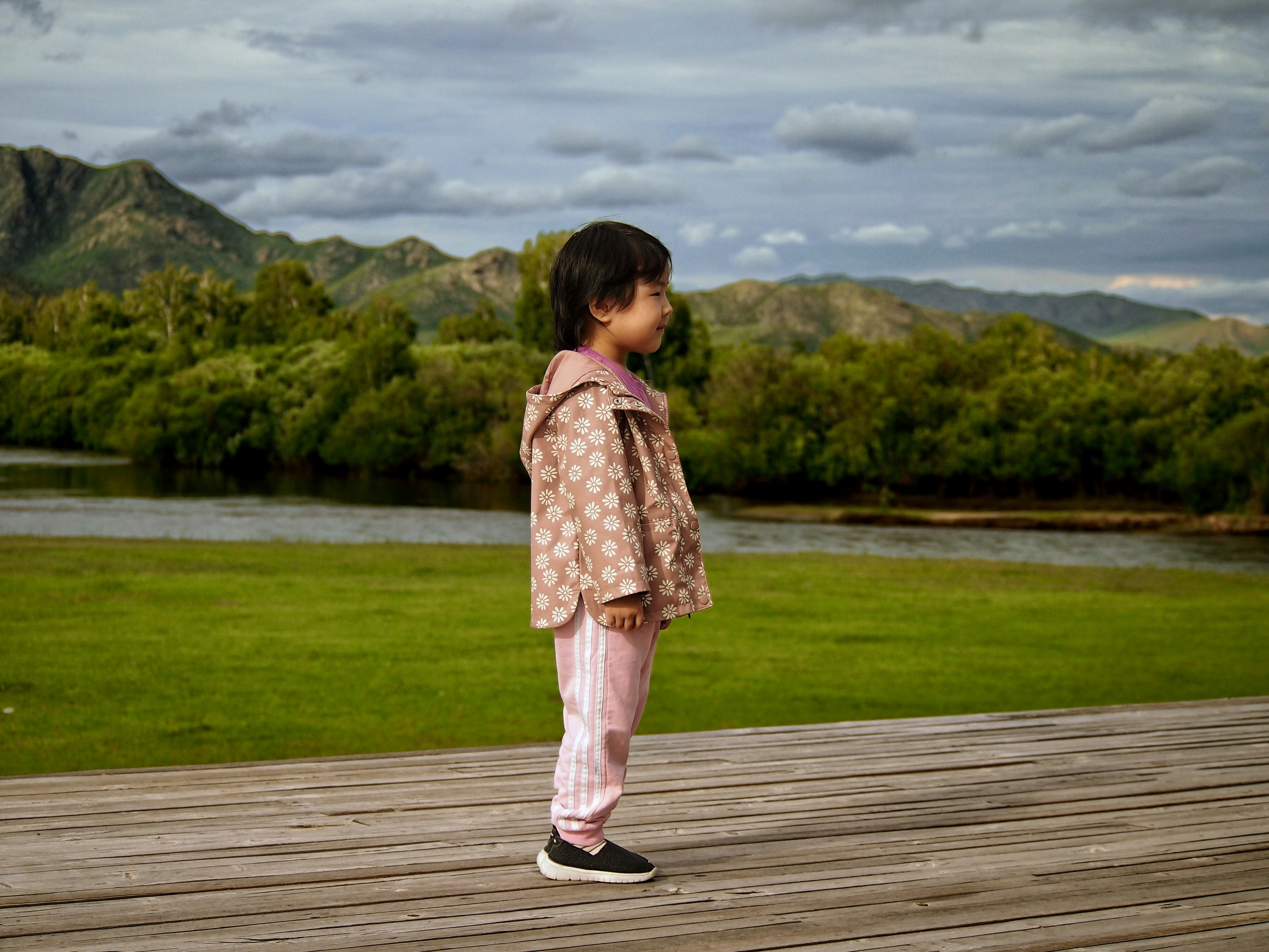 Young child in a patterned pink jacket and pink pants stands on a wooden deck by a river, with mountains and trees in the distance.