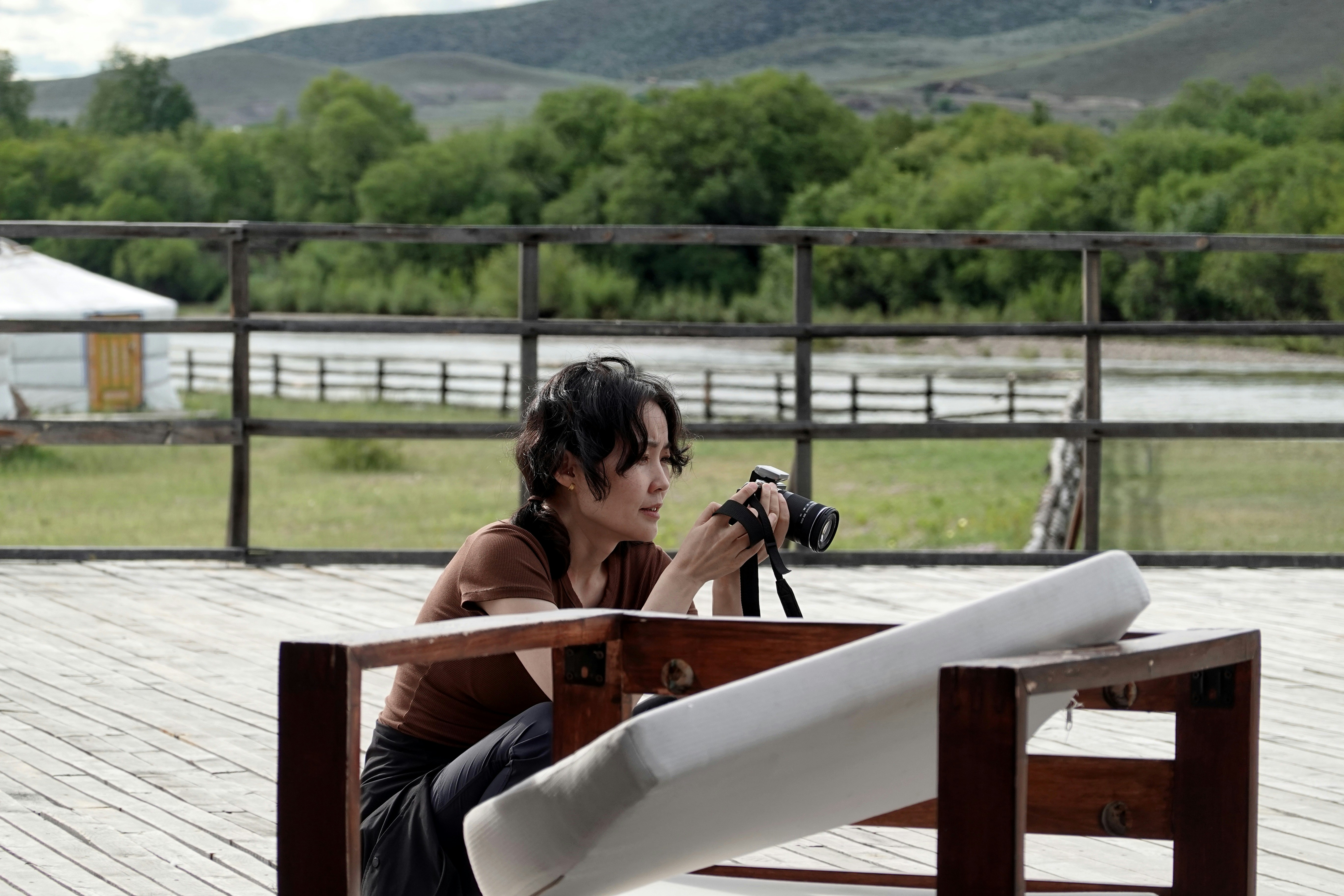 A photographer crouches at a wooden dock, framing a white kayak-like object with a camera. The scene conveys intent capture in a calm outdoor setting.