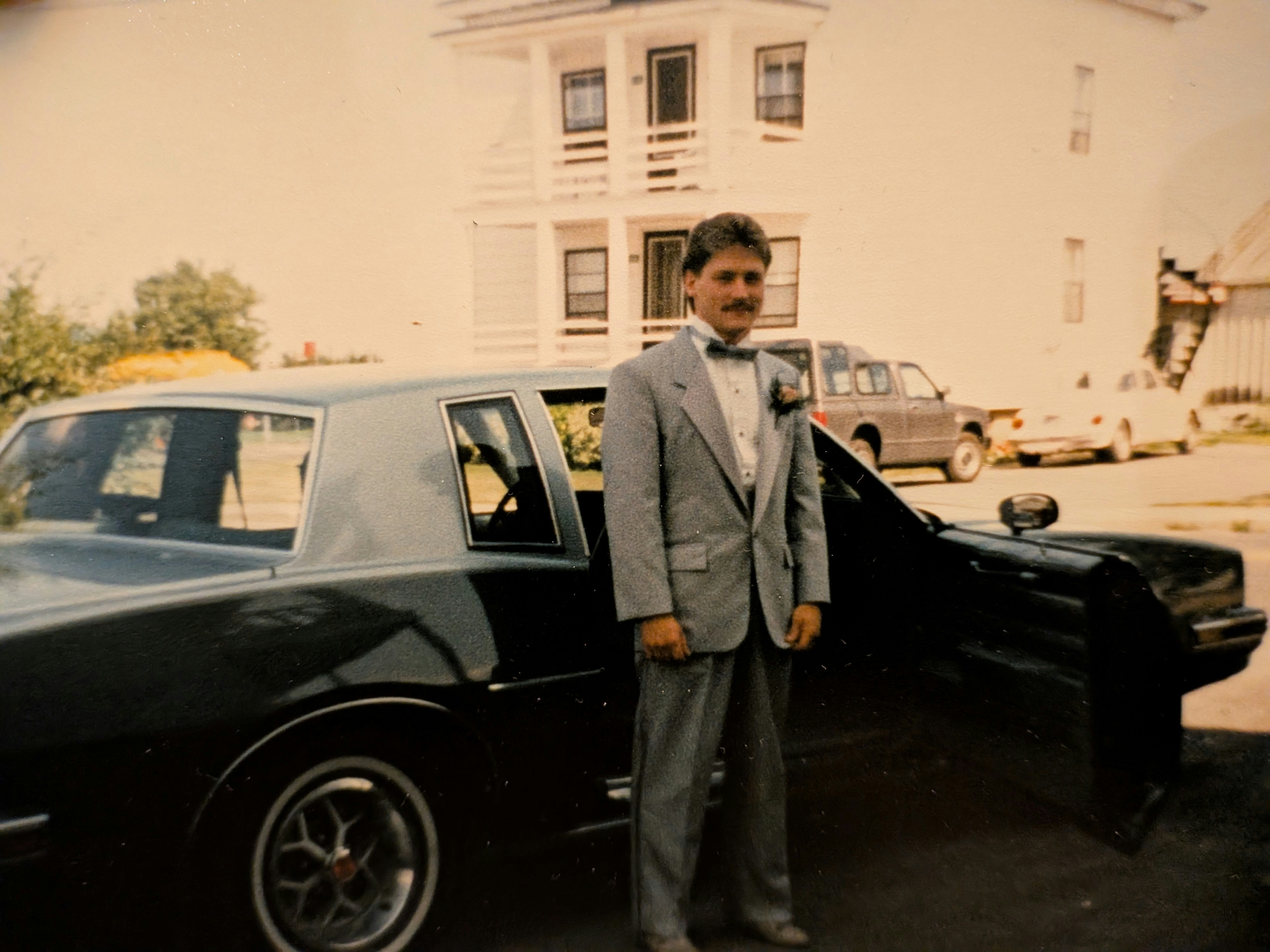A man in a light gray suit stands beside a vintage dark car in front of a light-colored building with several windows; the scene evokes a nostalgic, formal moment. This is a photographed scene with a classic car and attire.