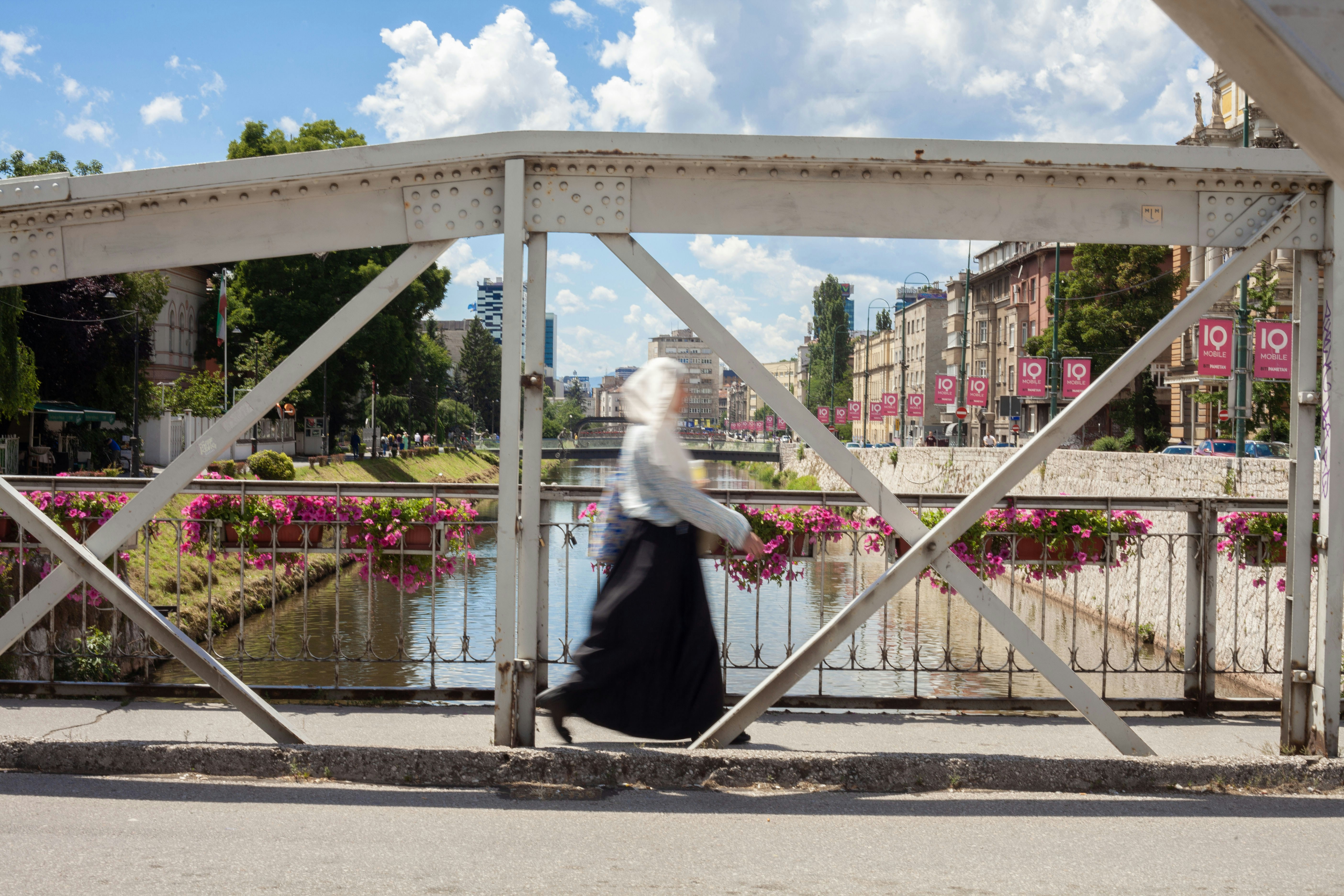 A blurred figure walks across a metal truss bridge over a canal, city buildings and flowers lining the background. The motion blur suggests a fleeting moment during a daytime stroll; the scene captures urban life with a decorative riverside promenade.