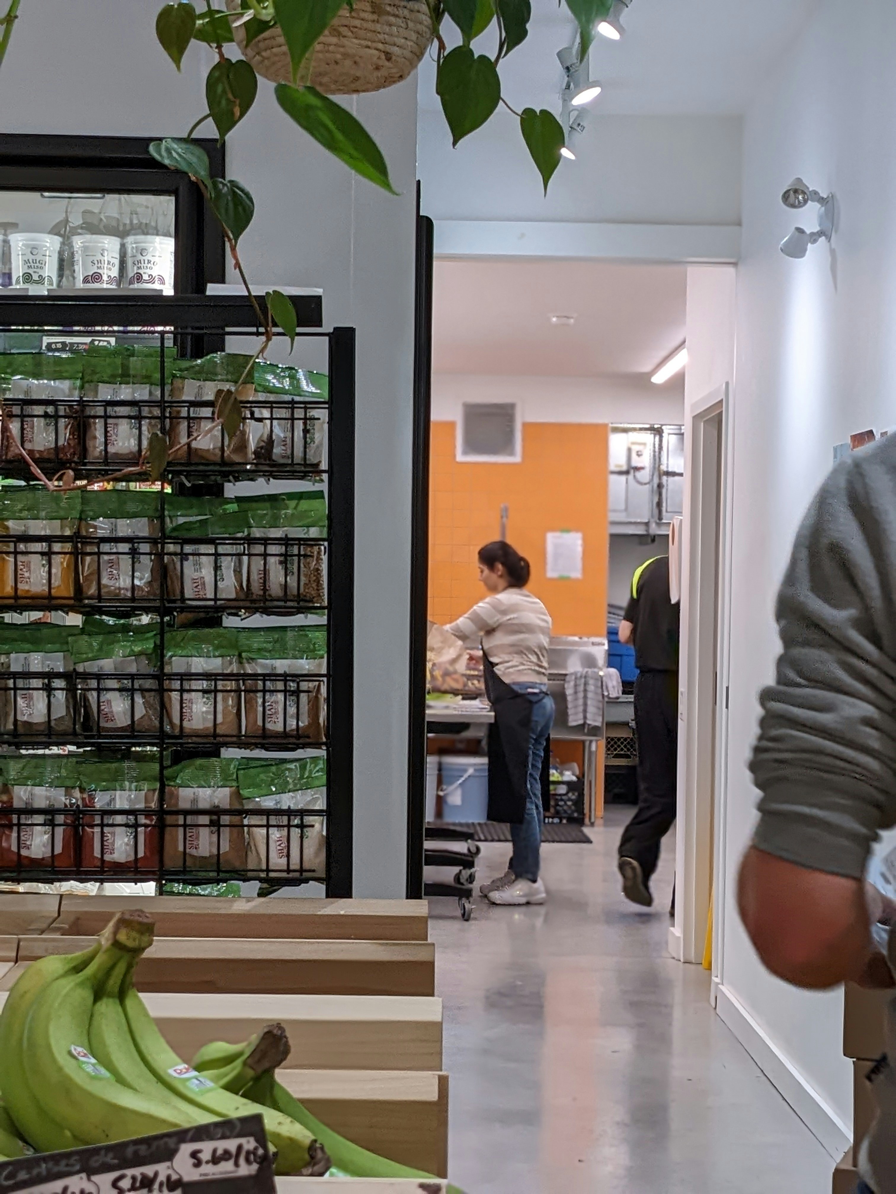 A shopper-friendly grocery store scene with a bright kitchen area in the background where a person stands by a counter, bananas in the foreground, and another partial figure to the right. The setting suggests a casual workflow space within the store.