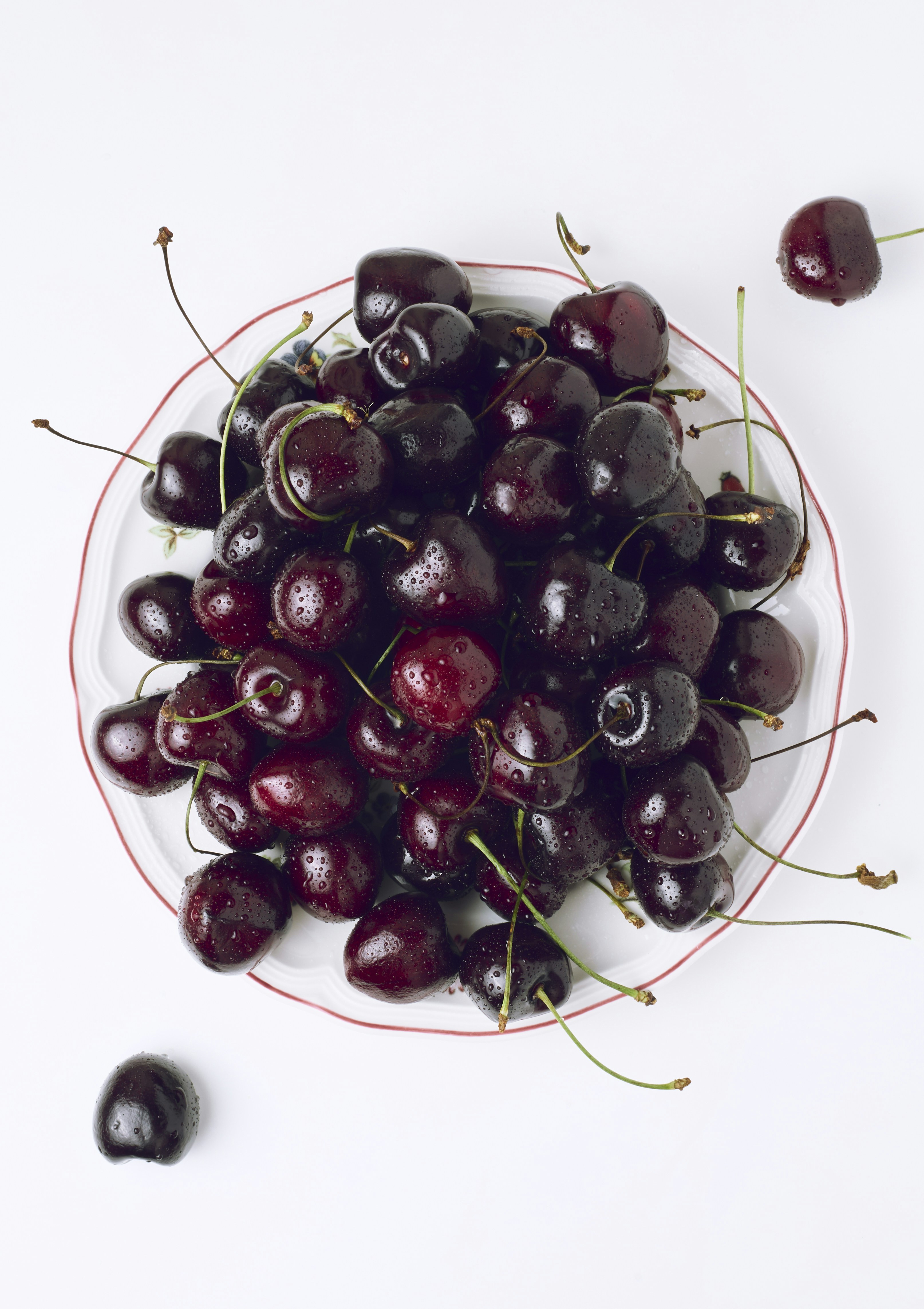 A bowl of dark, dewy cherries sits on a light surface, with one cherry stray at the bottom left; the scene highlights fresh fruit with close, high-contrast detail.
