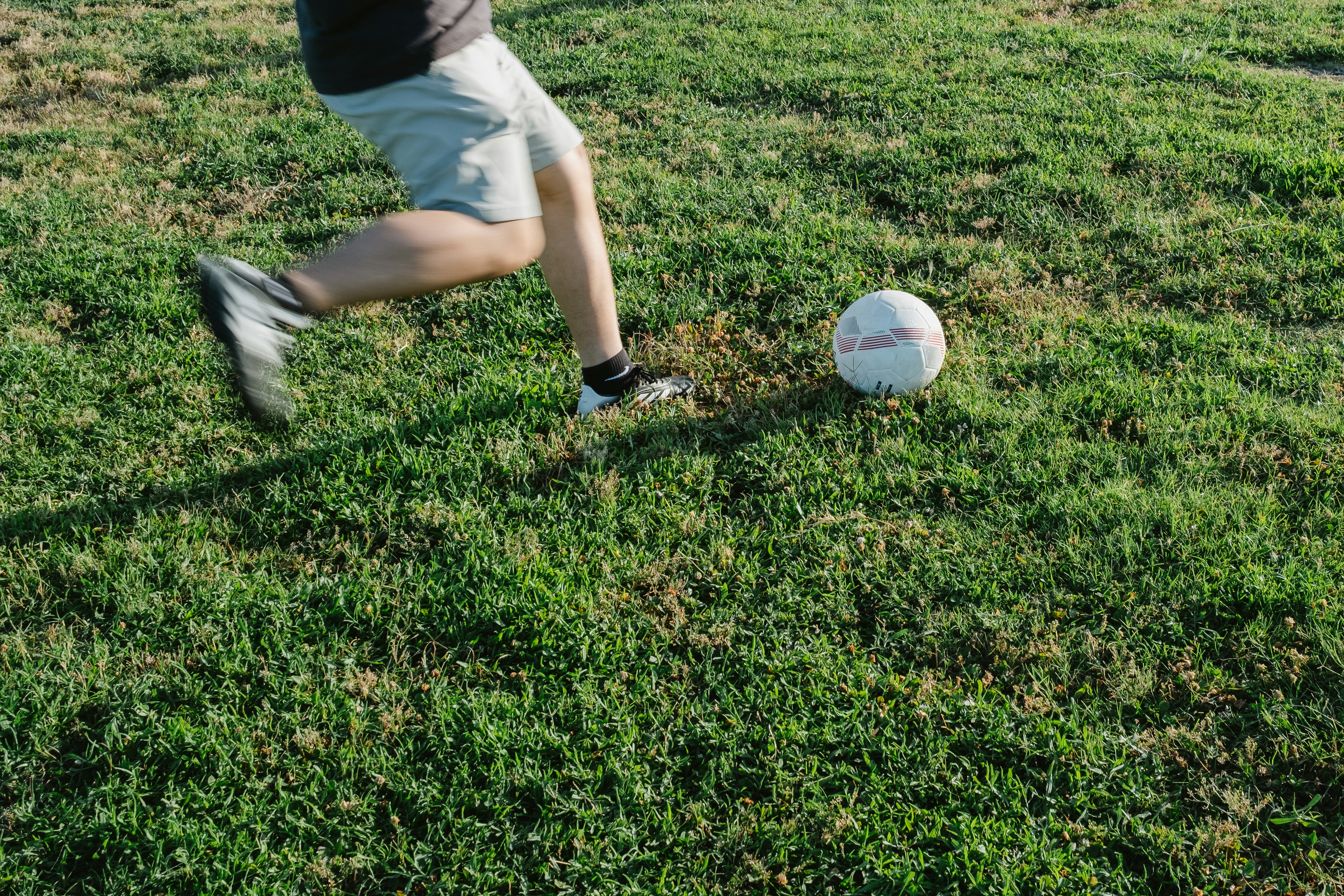 A young player charges across a sunlit grassy field toward a weathered soccer ball, capturing a brief, dynamic moment of motion and play.