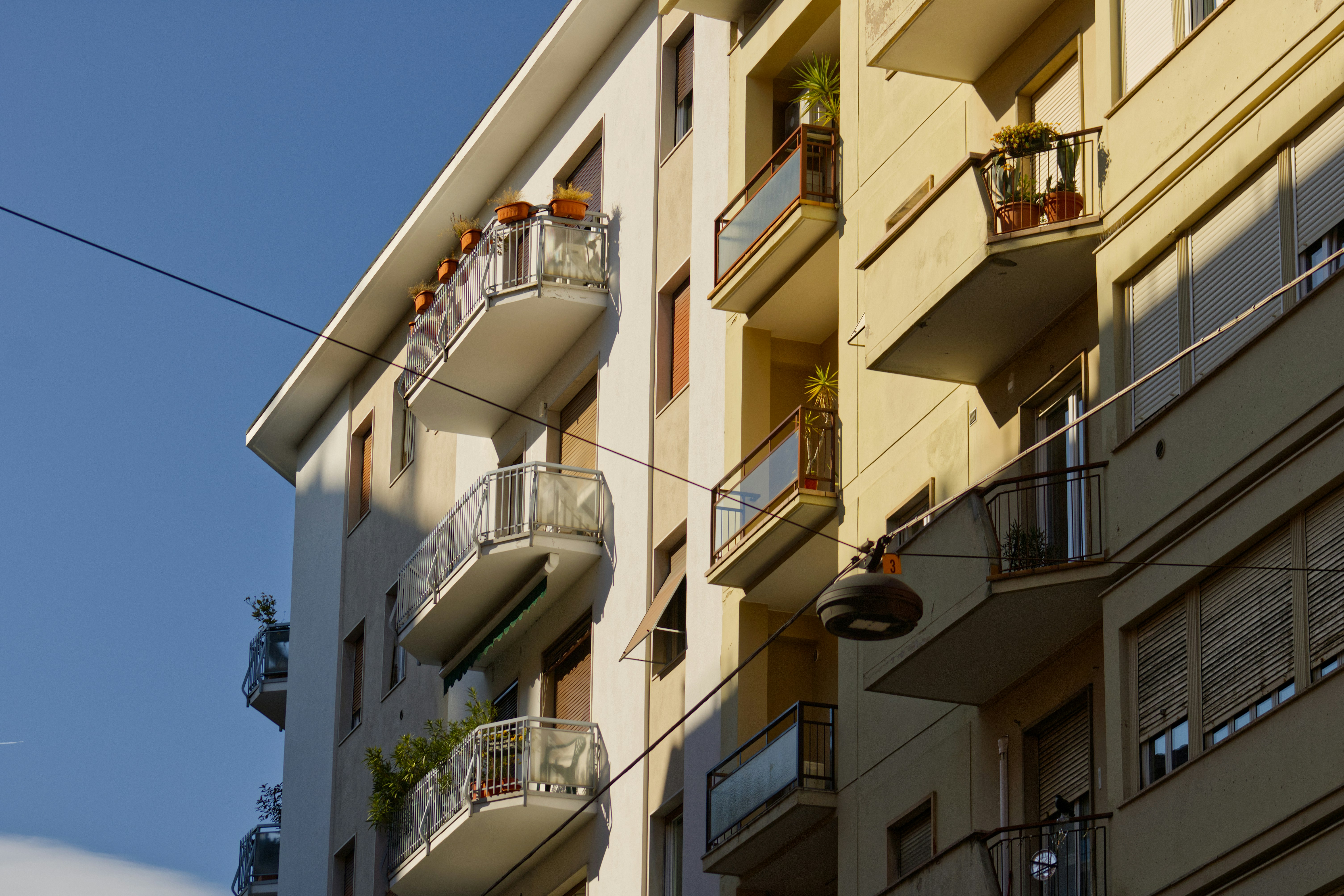 Rows of balconies on a sunlit apartment building, with potted plants and a single street lamp against a blue sky.
