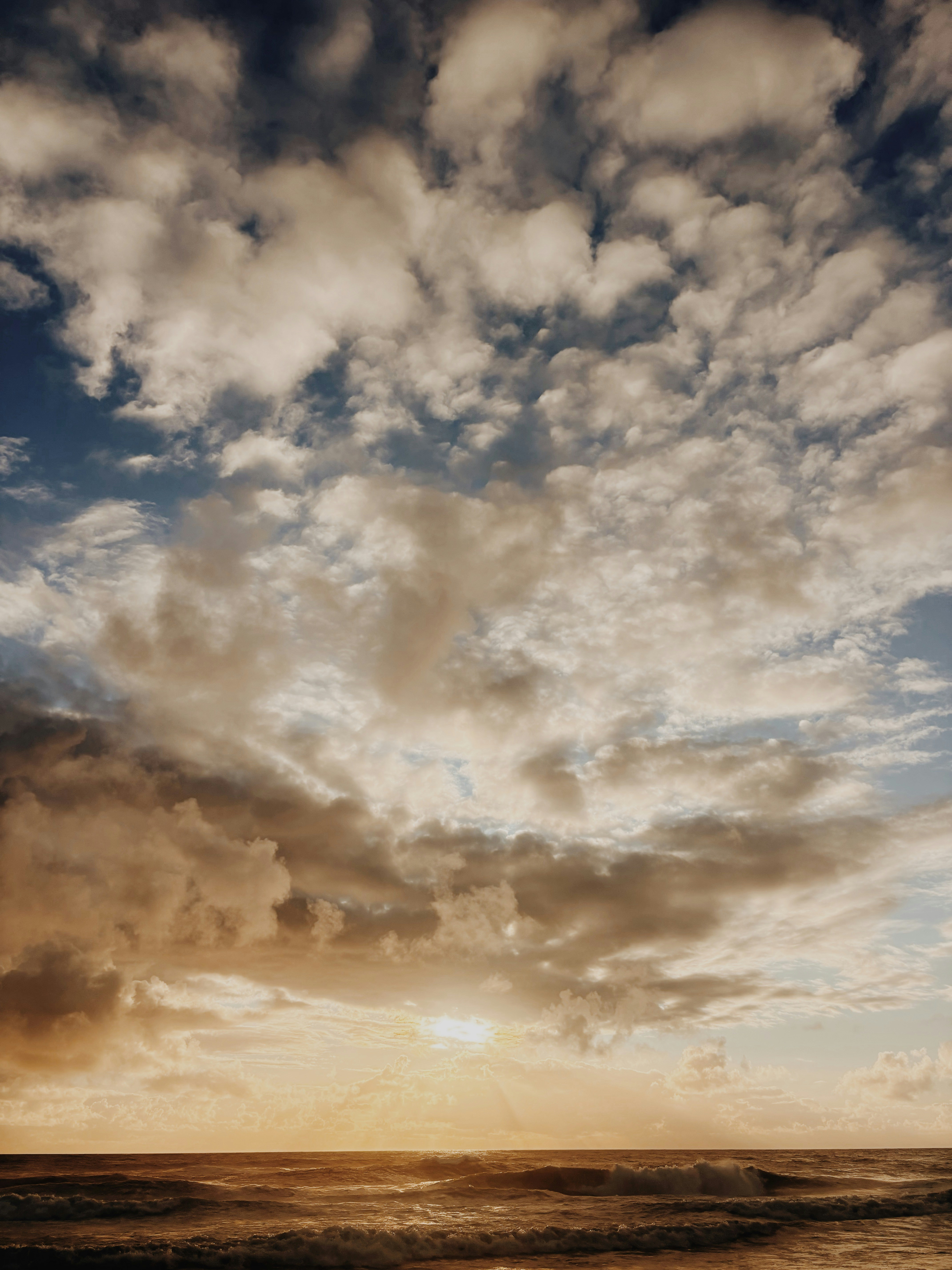 Warm sunset over a calm sea with dramatic clouds filling the sky; horizon line anchors the scene and suggests a serene, expansive atmosphere. Photograph of a seascape showing light breaking through clouds near the horizon.