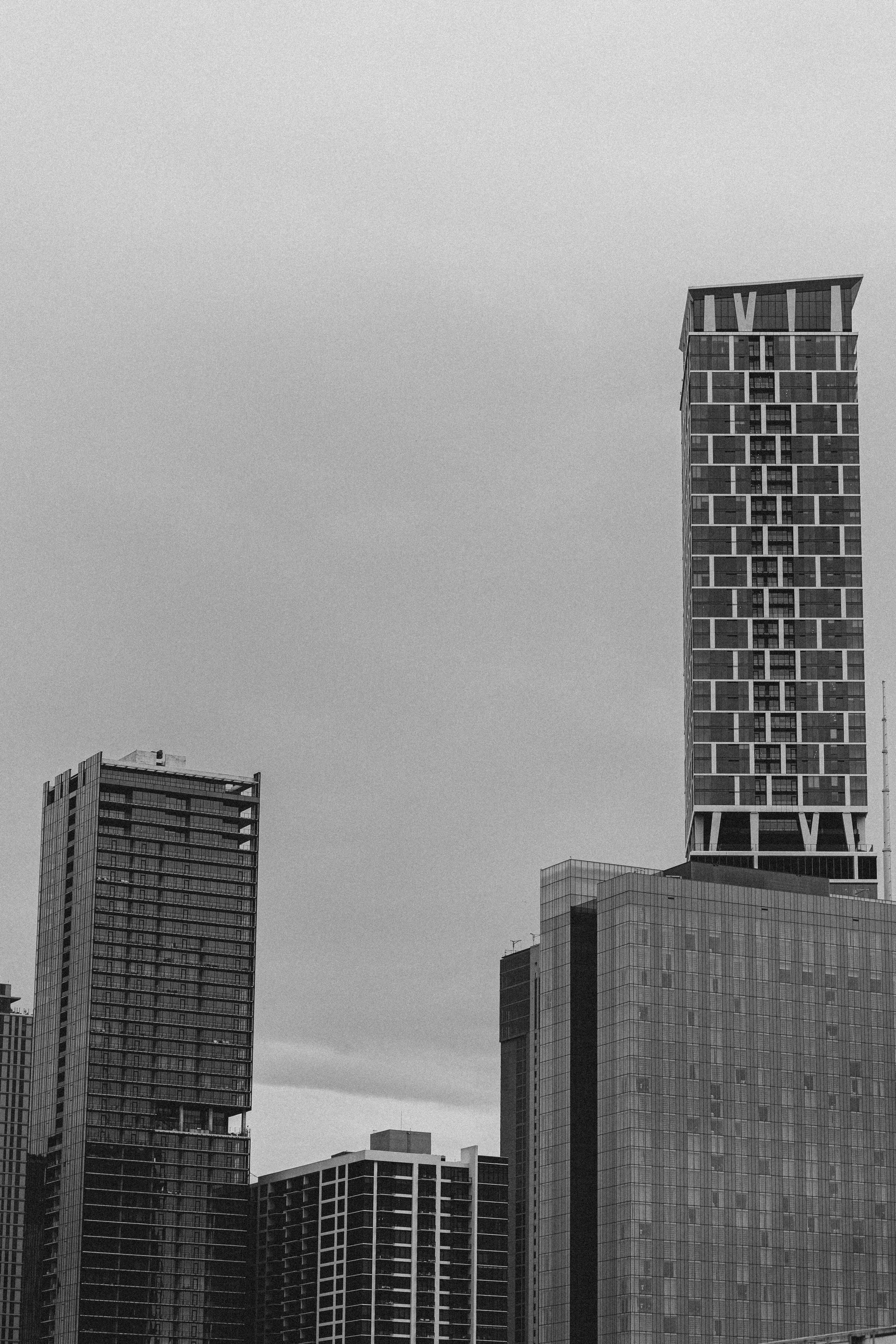 Monochrome cityscape with tall, modern buildings rising against a cloudy sky, emphasizing architectural geometry and depth.