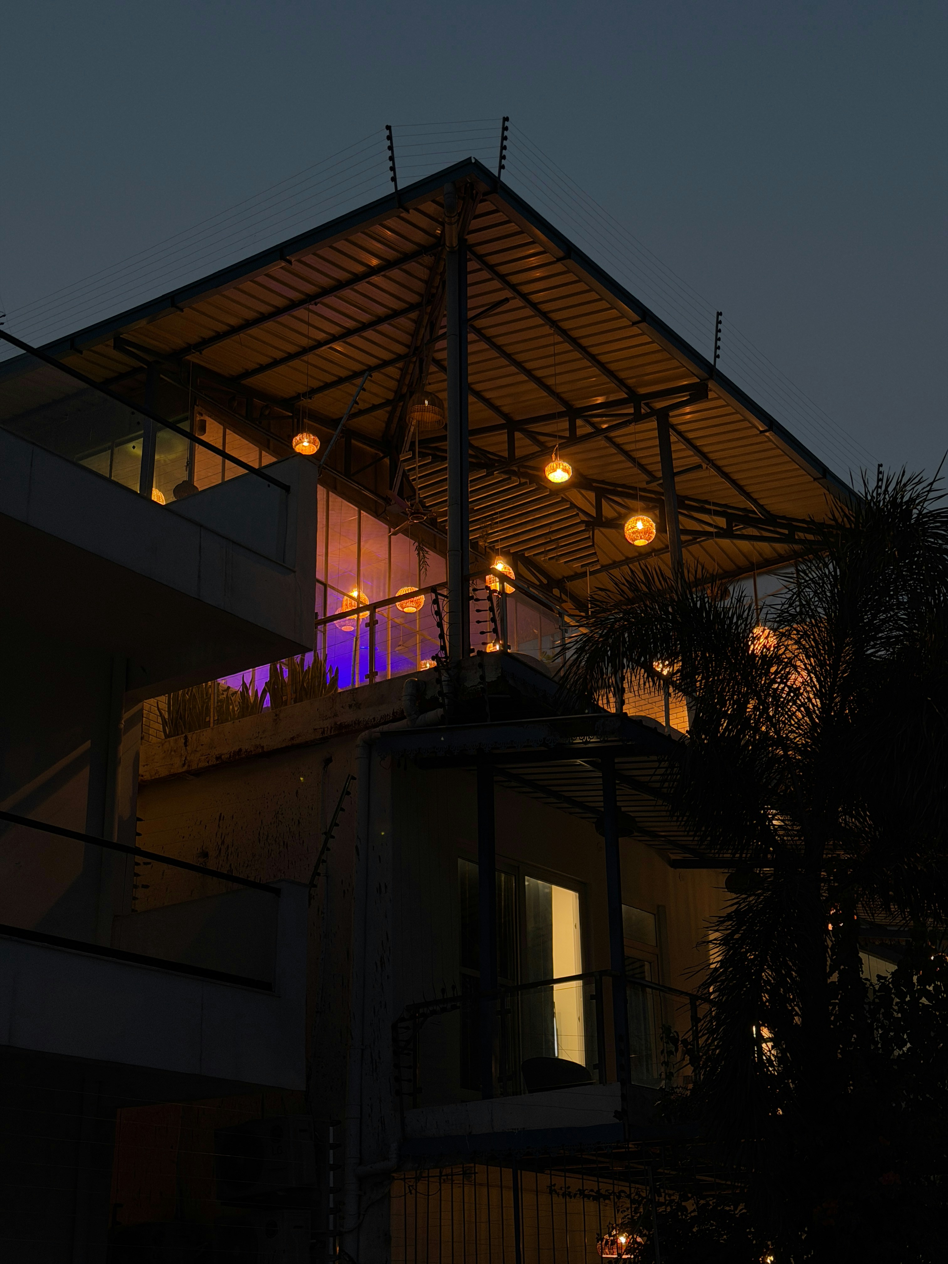 Night scene of a modern multi-level terrace building lit by warm bulbs, with glass walls and a hint of purple light in the interior.