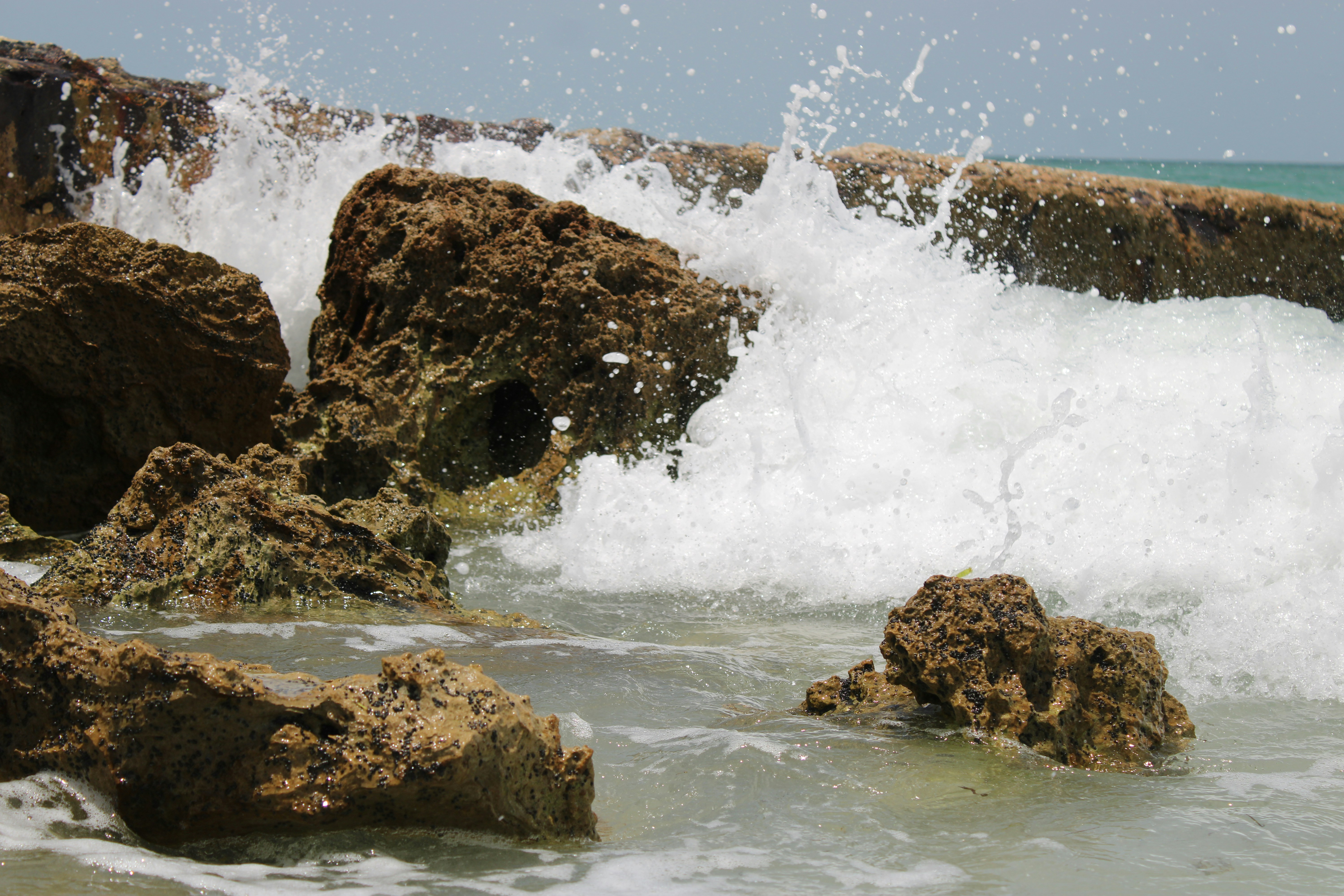 Waves crash over rugged rocks along a sunlit shoreline, creating white spray and wet surges against a calm horizon.