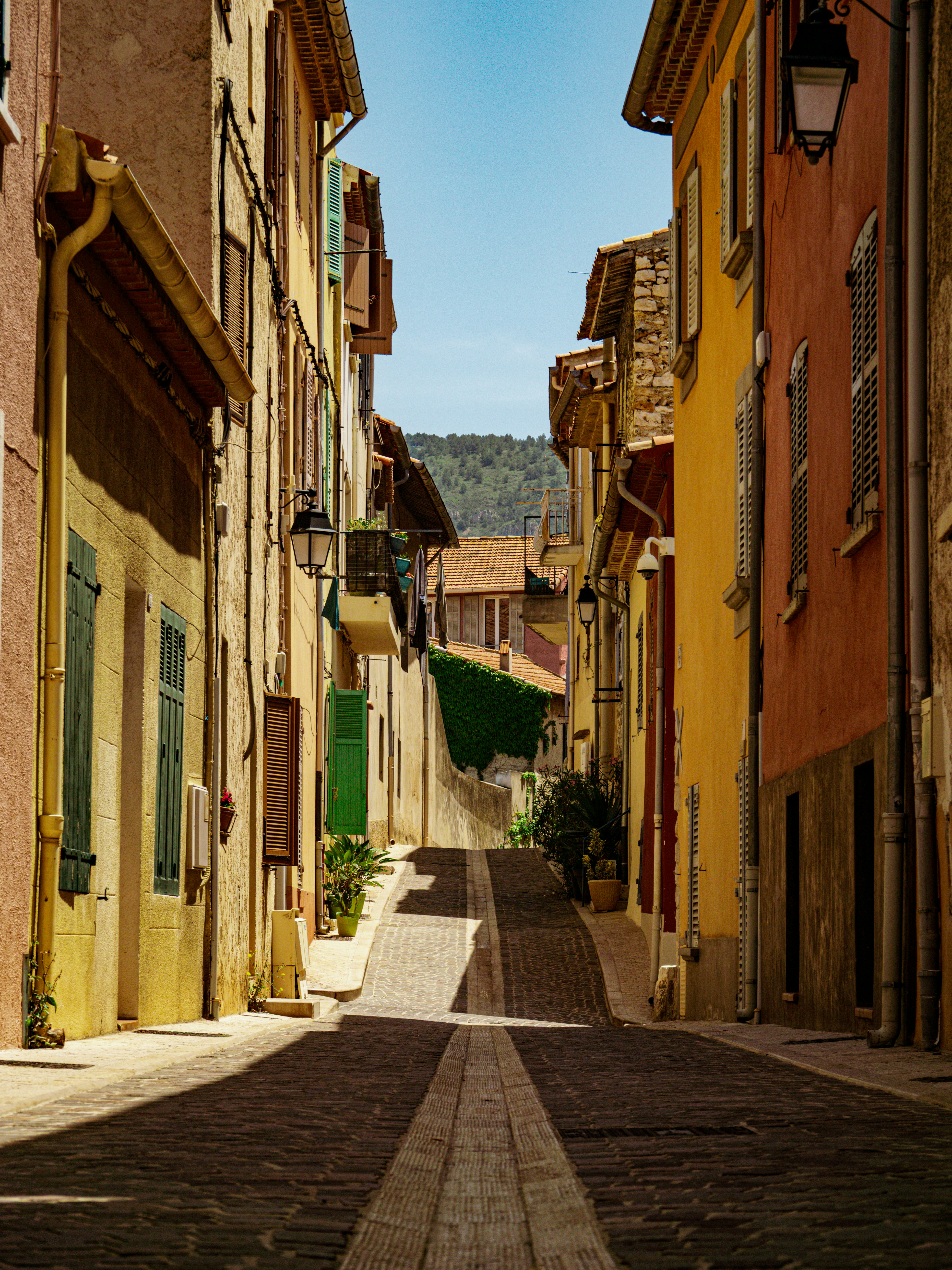 Sunlit cobbled street in a colorful hillside village, with narrow buildings and shuttered windows lining a central path toward a distant hill.