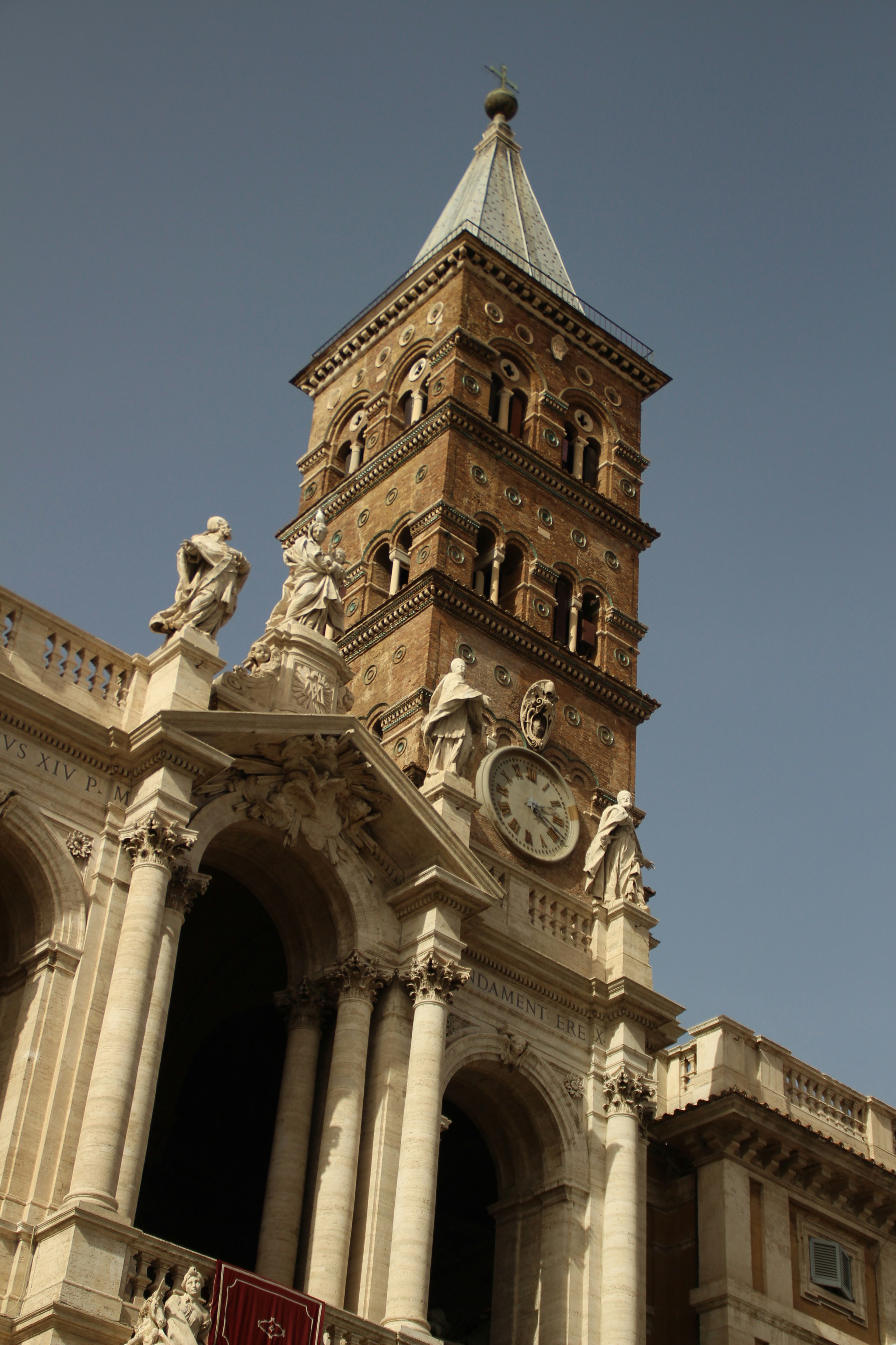 Stone archway and ornate statues frame a tall brick bell tower with a clock, set under a pale blue sky; architectural details and shadows highlight the historic facade.