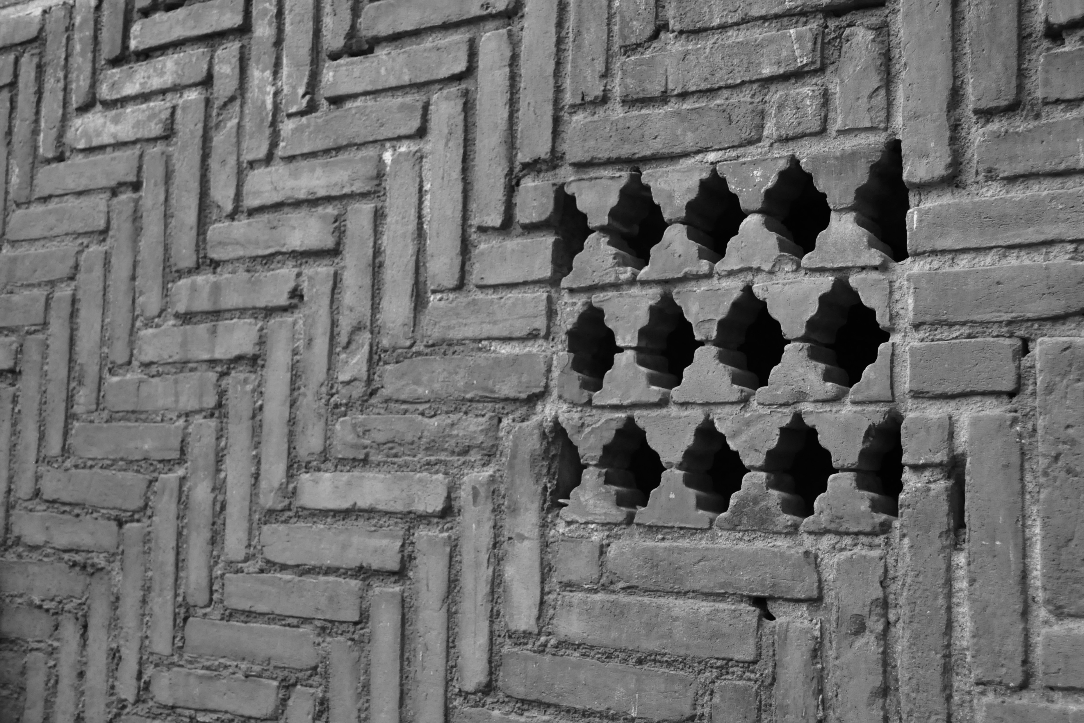 Textured brickwork with diagonal chevron patterns and a perforated vent cluster as the focal point. Concrete tones and subtle light yield a quiet architectural detail.