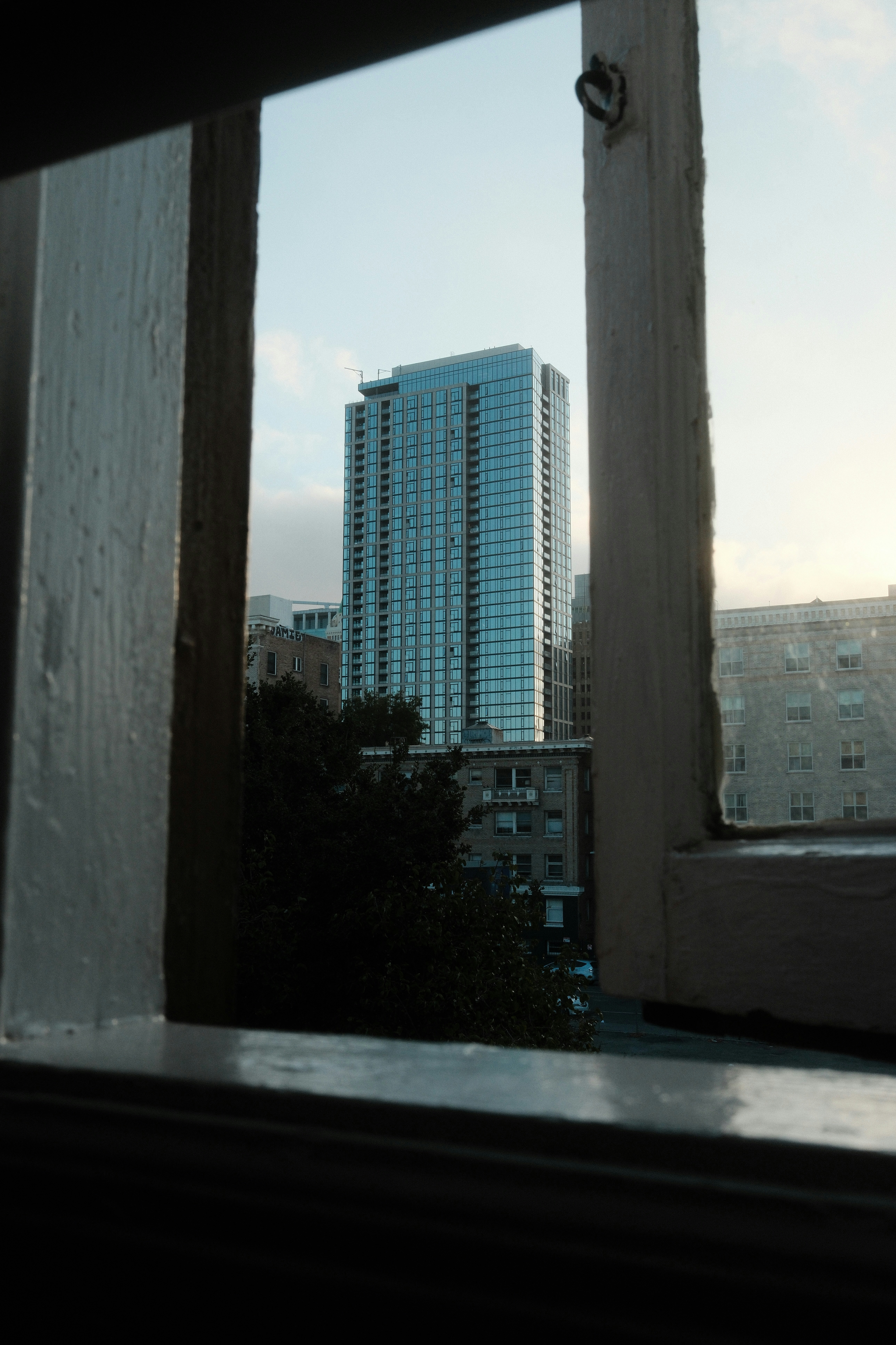 Urban skyline framed by an old window, with a tall glass building rising above surrounding structures.