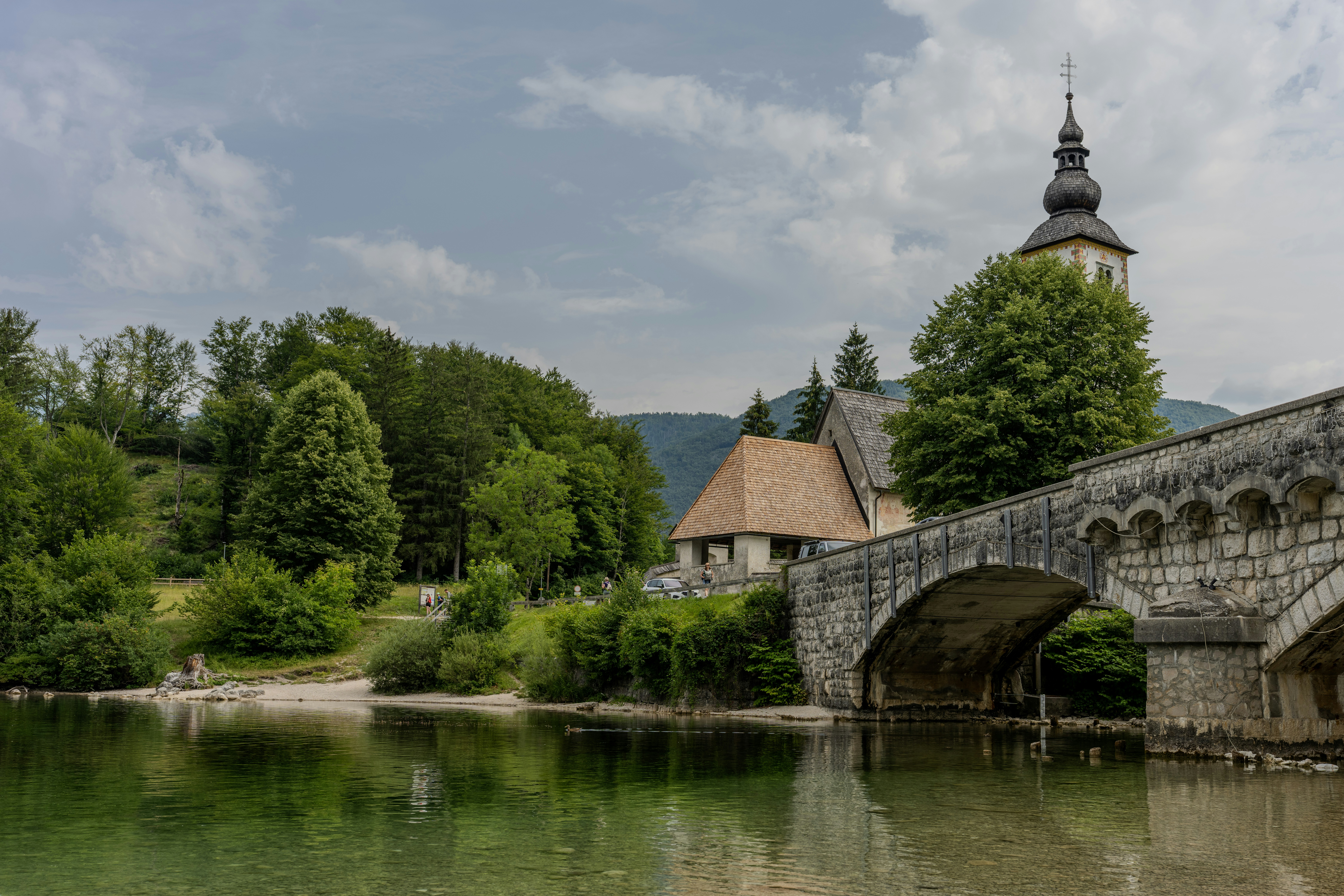 Scenic riverside with a stone bridge, historic buildings, and lush trees along calm water, highlighting rustic architecture and tranquil landscape.