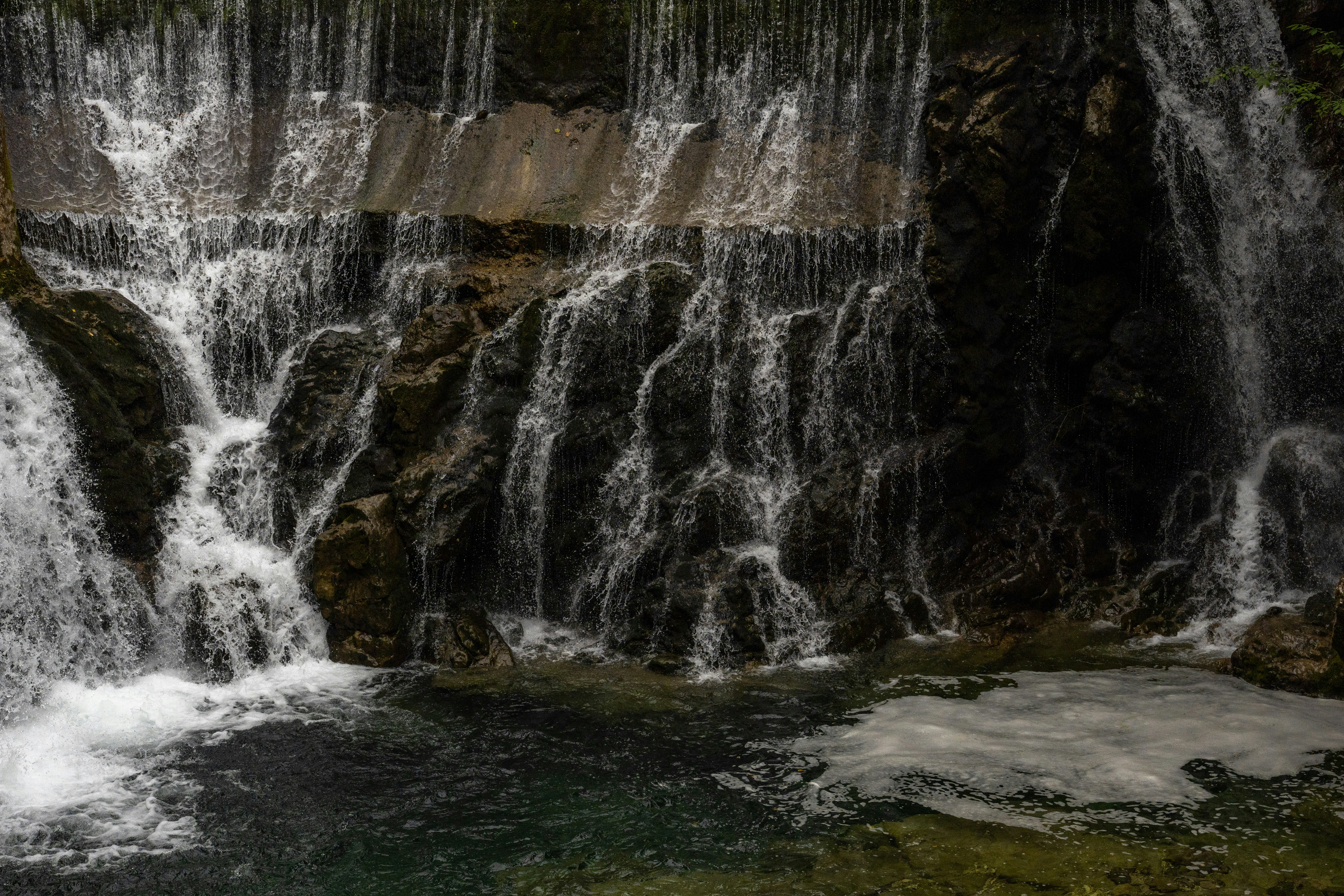 A multi-tiered waterfall cascades over dark rocks into a pool.