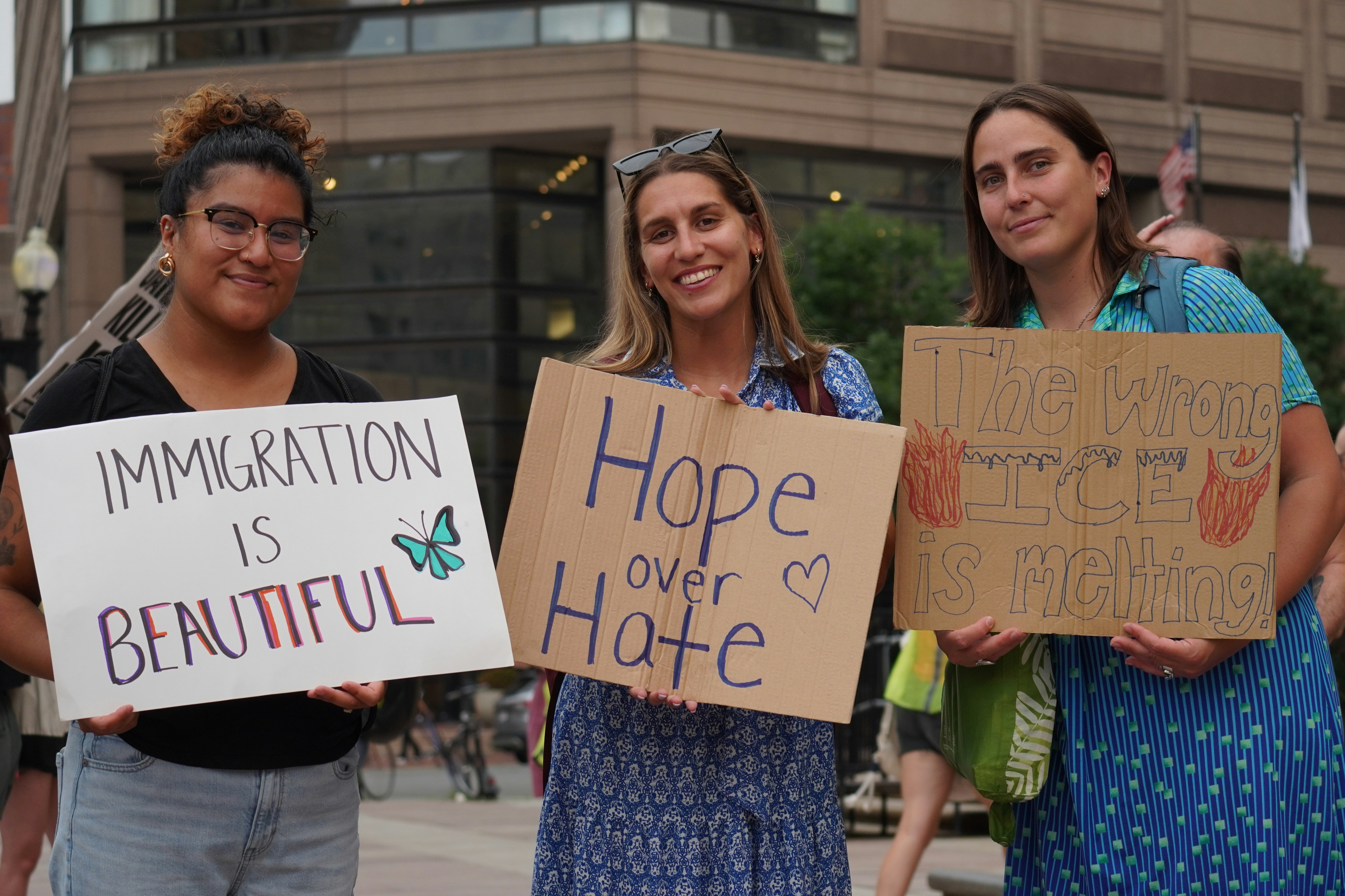Three women hold protest signs against intolerance in an urban setting, conveying messages of immigration pride, anti-hate, and resilience.