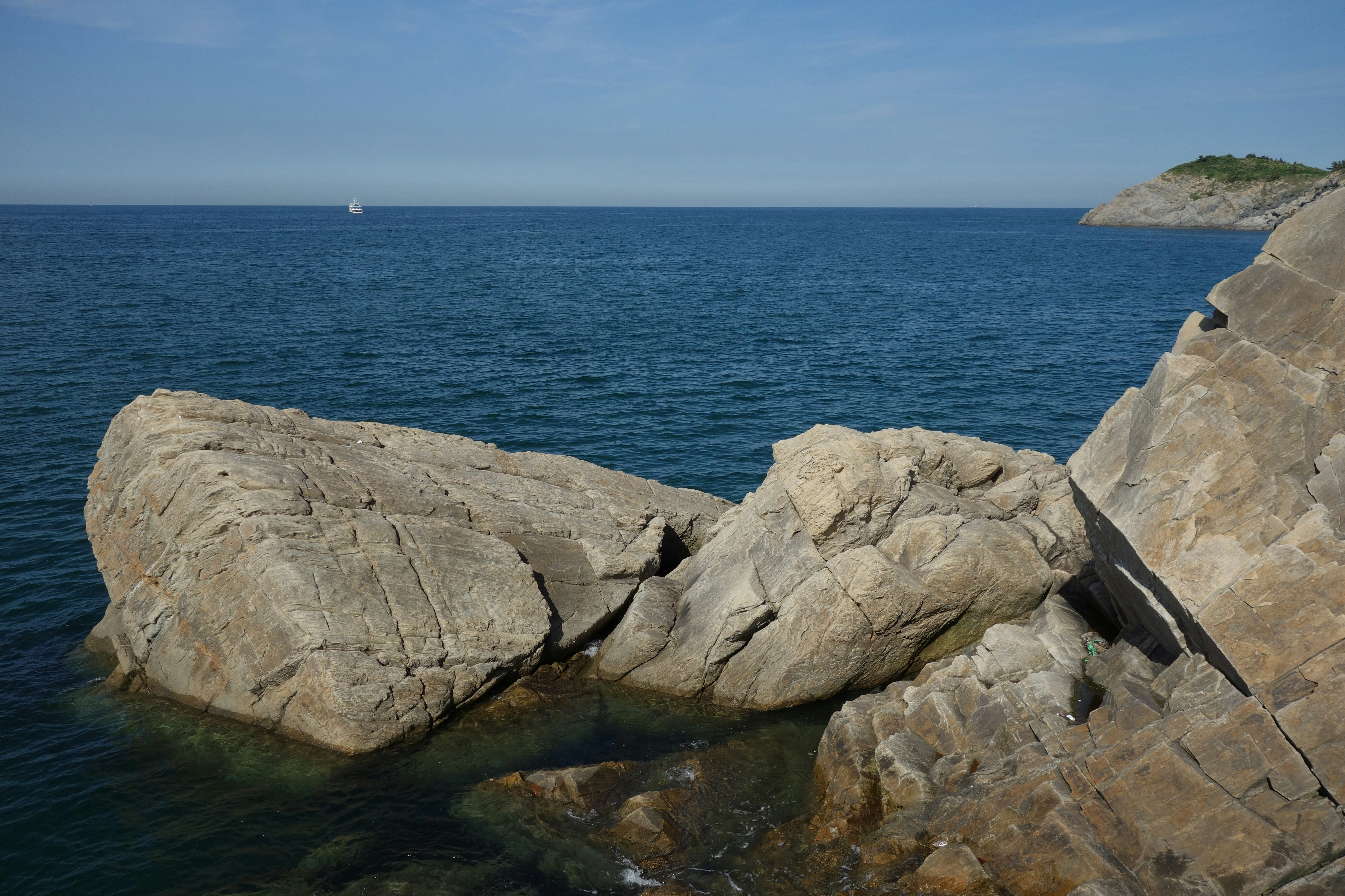 Sunlit rocky shoreline with calm blue sea stretching to the horizon; rugged stone formations in the foreground anchor the scene. This is a natural seascape photograph.
