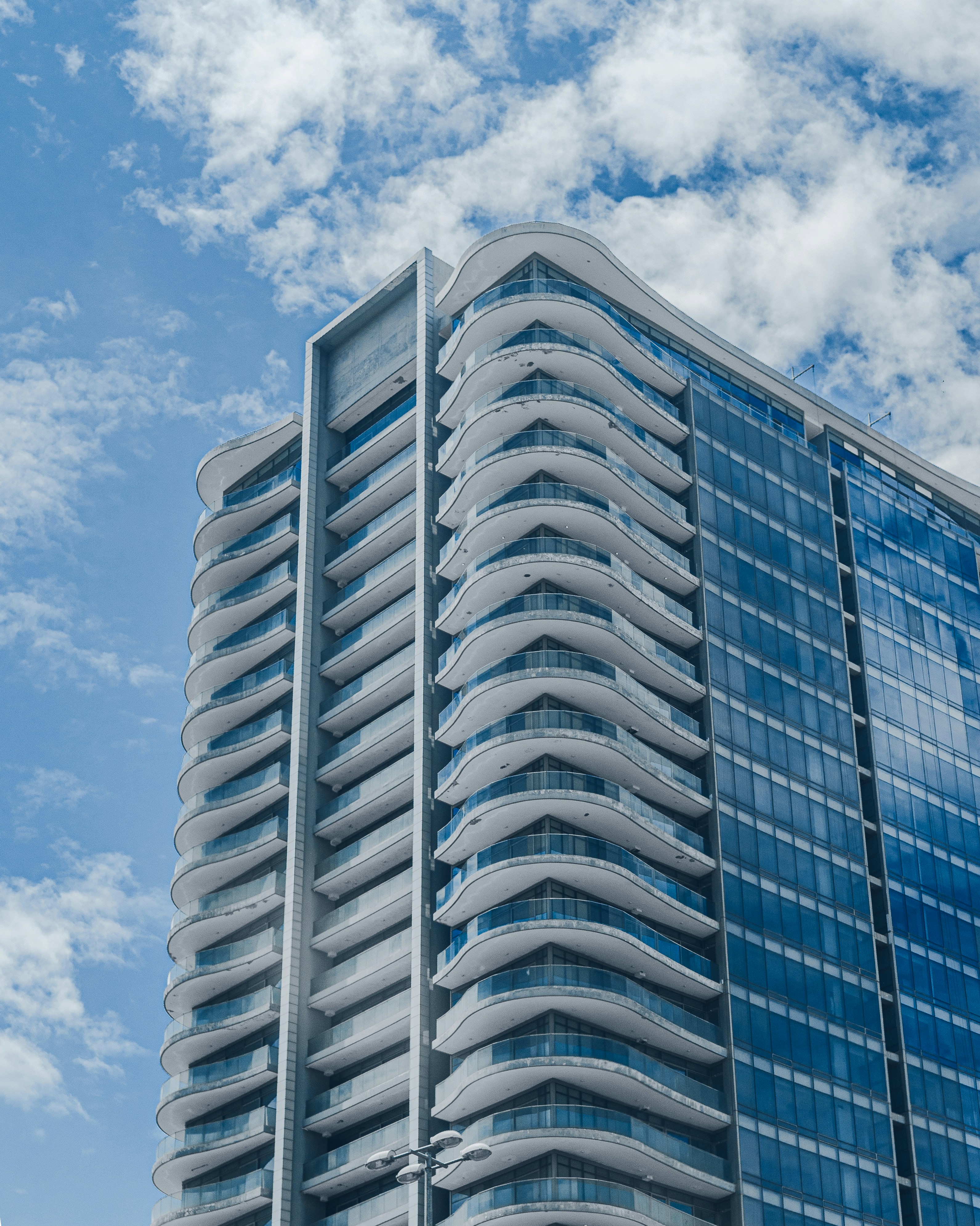 Tall glassy building with repeating curved balconies against a blue sky and scattered clouds; architectural portrait emphasizing vertical rhythm.