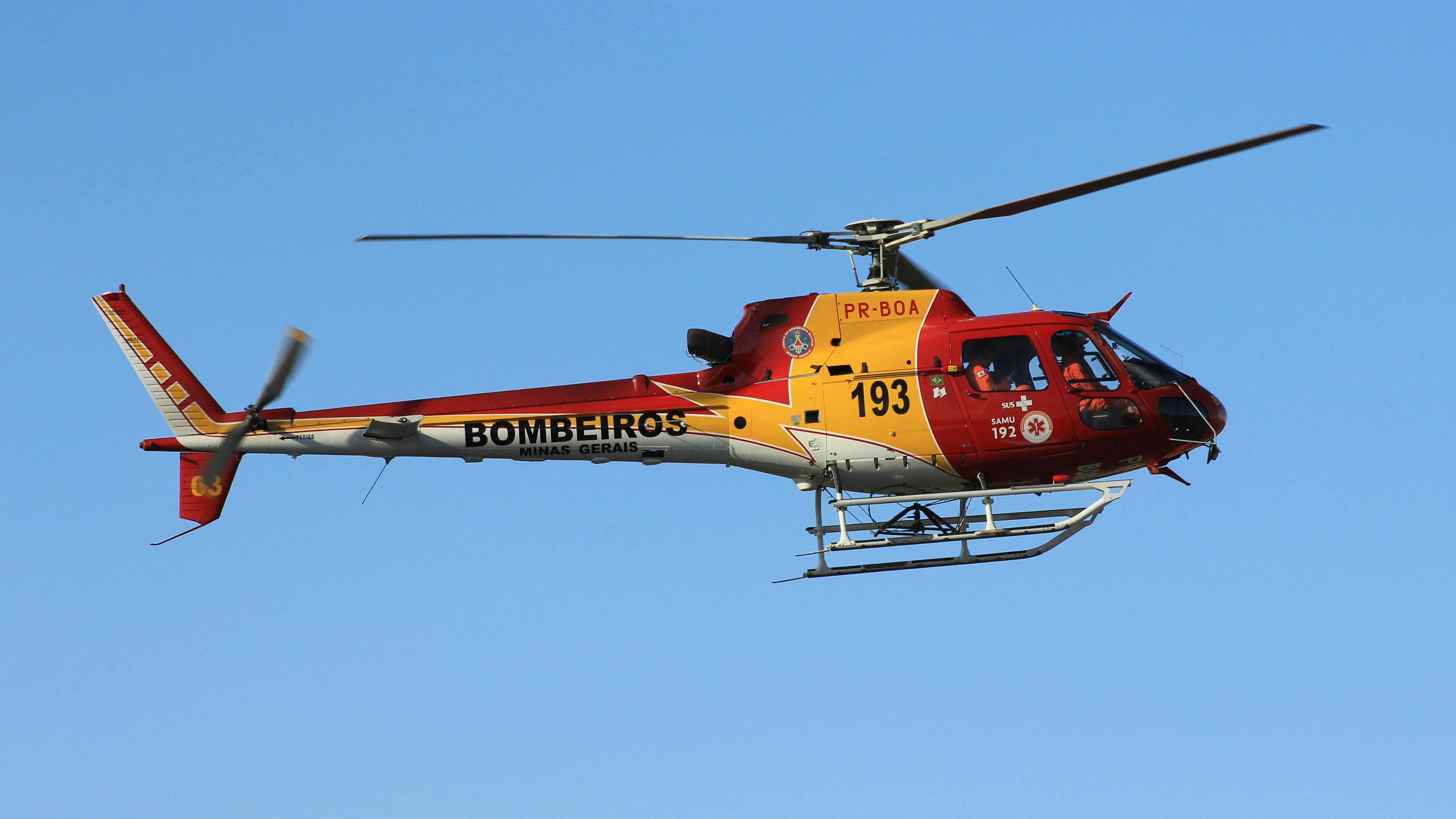 Bright red and yellow firefighting helicopter in mid-flight against a blue sky, rotor blur indicating motion.