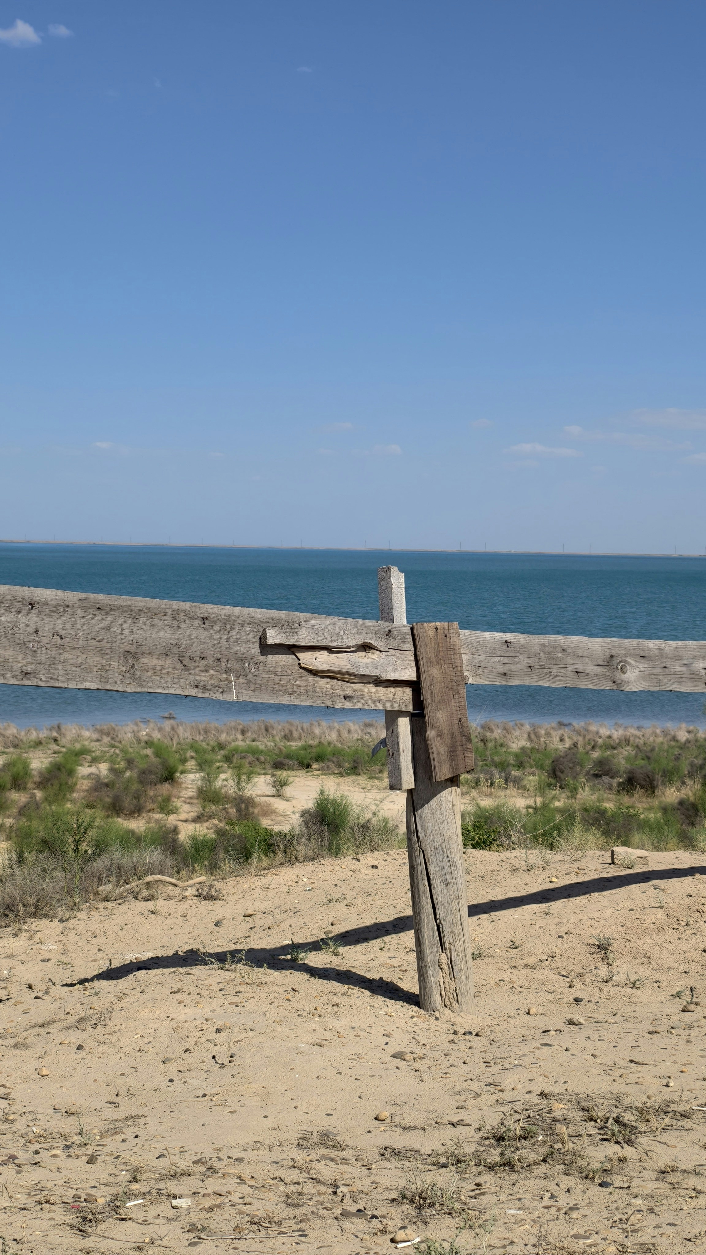 Rugged wooden fence stretches across a sandy coastal foreground with a calm blue sea and clear sky beyond.