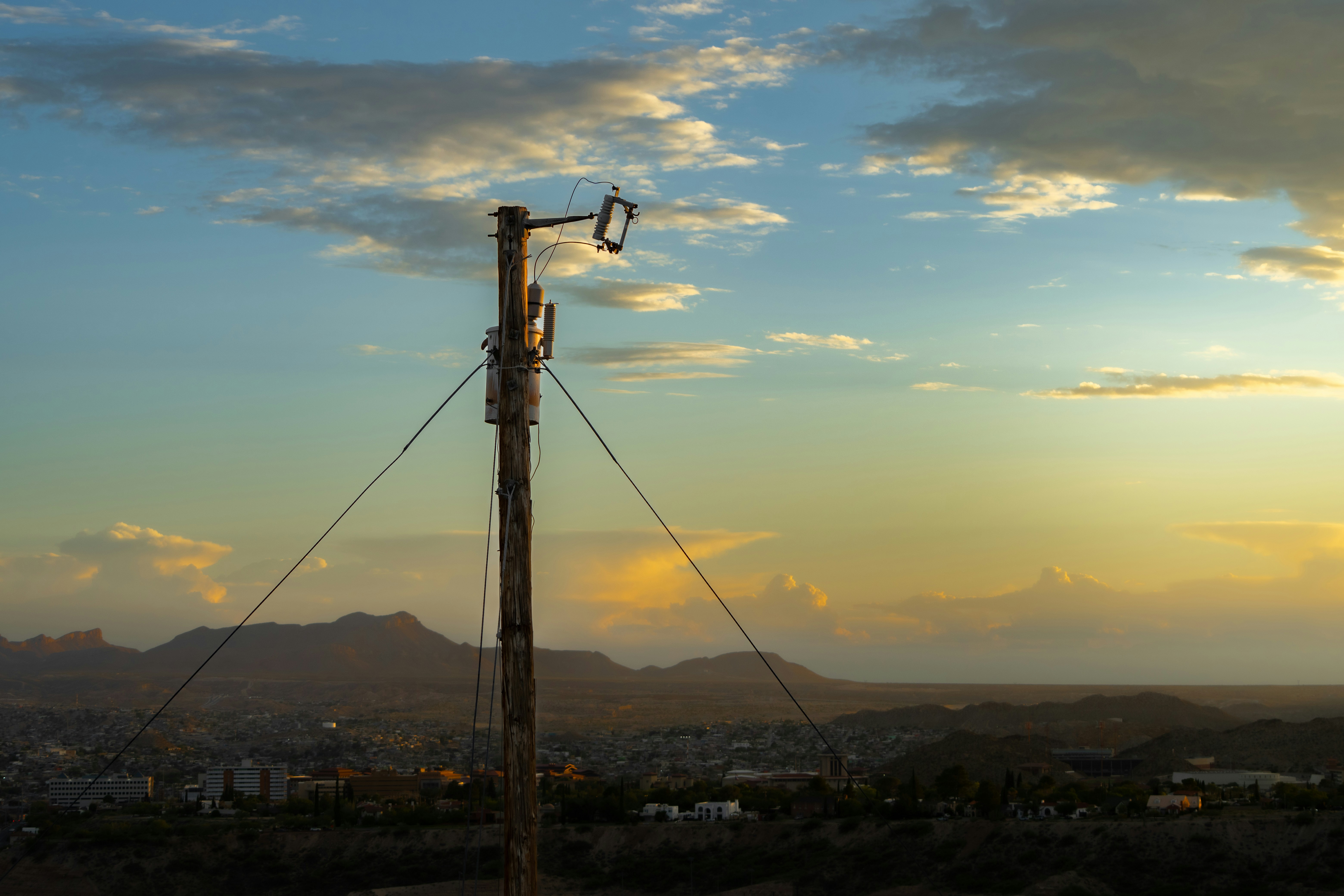 A solitary wooden utility pole with guy wires stands against a desert valley at sunset, silhouetted by a warm, expansive sky.