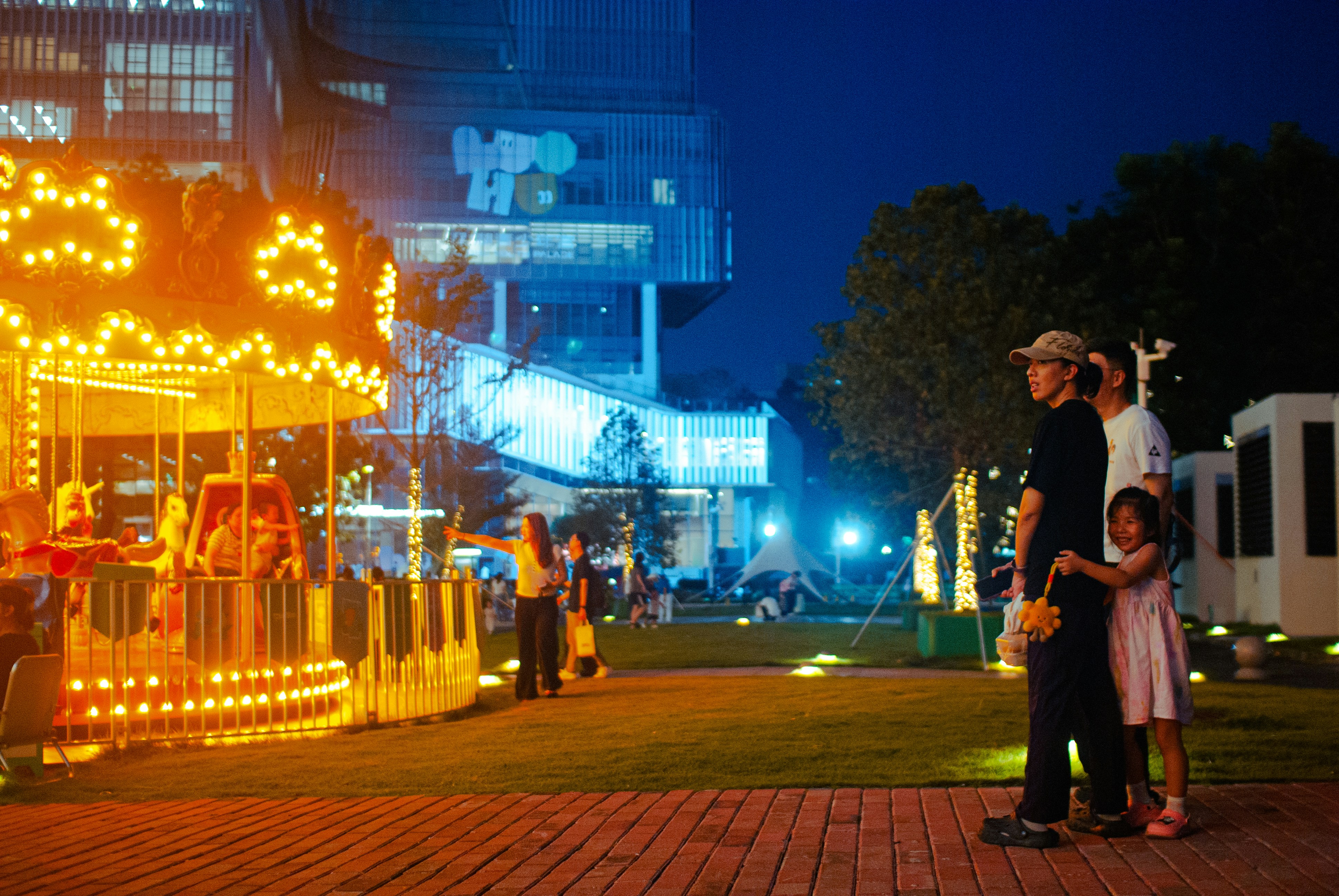 A night scene at a park with a brightly lit carousel on the left and a small family standing together on the right, illuminated by warm string lights and ambient blue city lights.