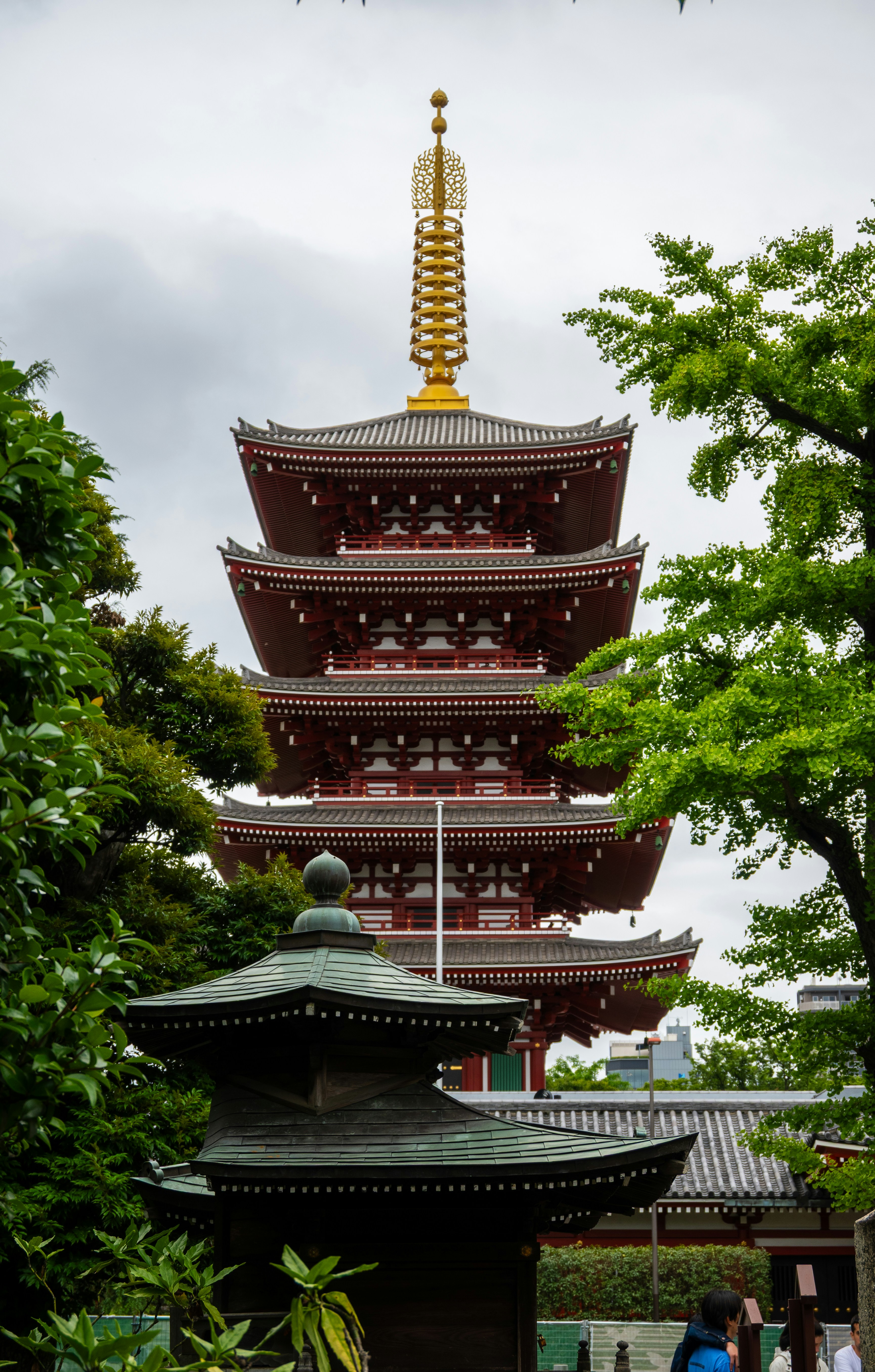 Multi-tiered red pagoda rising above lush greenery, with a smaller wooden structure in the foreground and an overcast sky in the background.