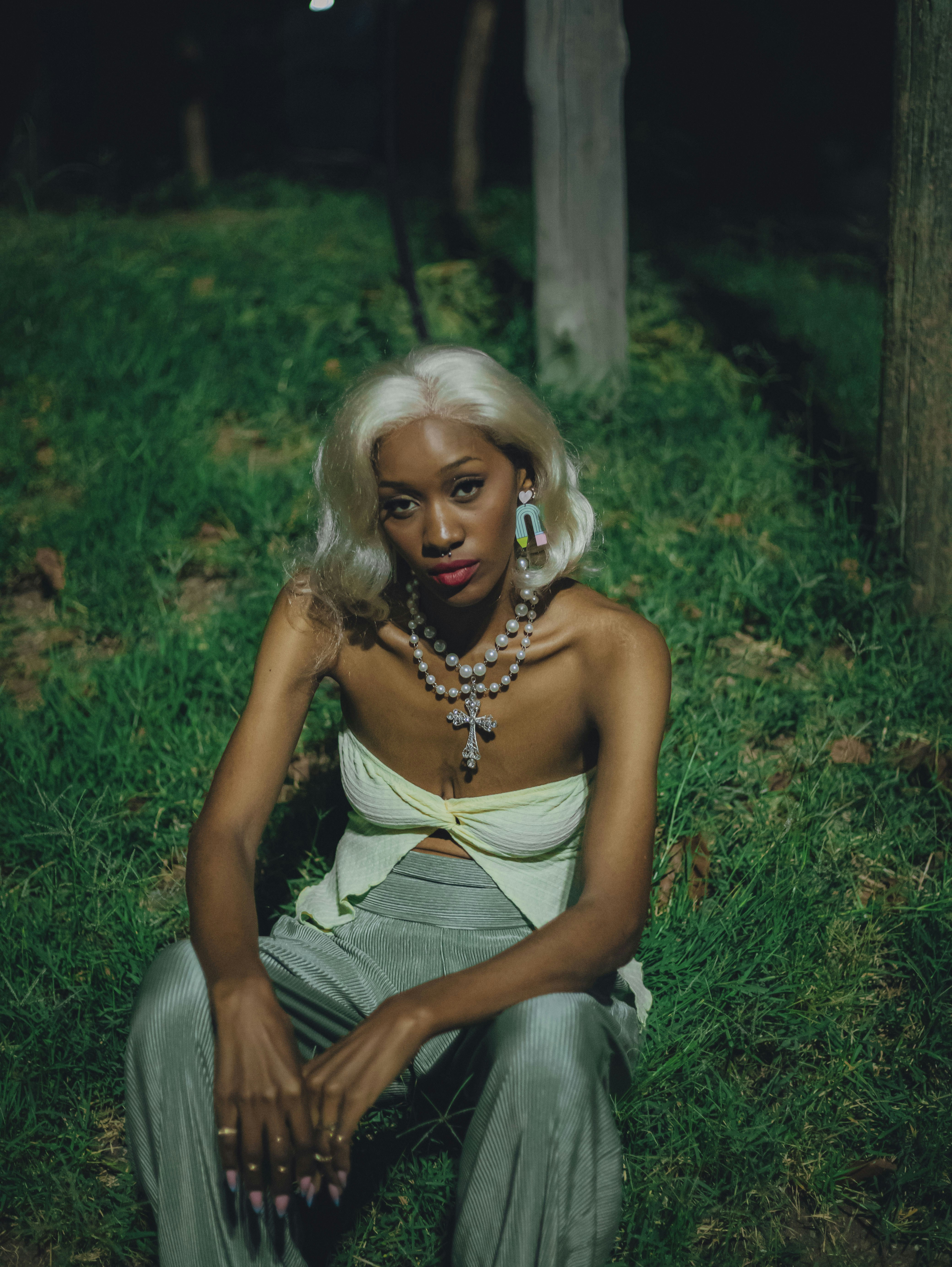 A poised woman with platinum blonde hair sits on grass at night, wearing layered pearl jewelry and a pale green wrap top. The scene emphasizes her calm, intimate presence in a dim, green-lit woodland setting.