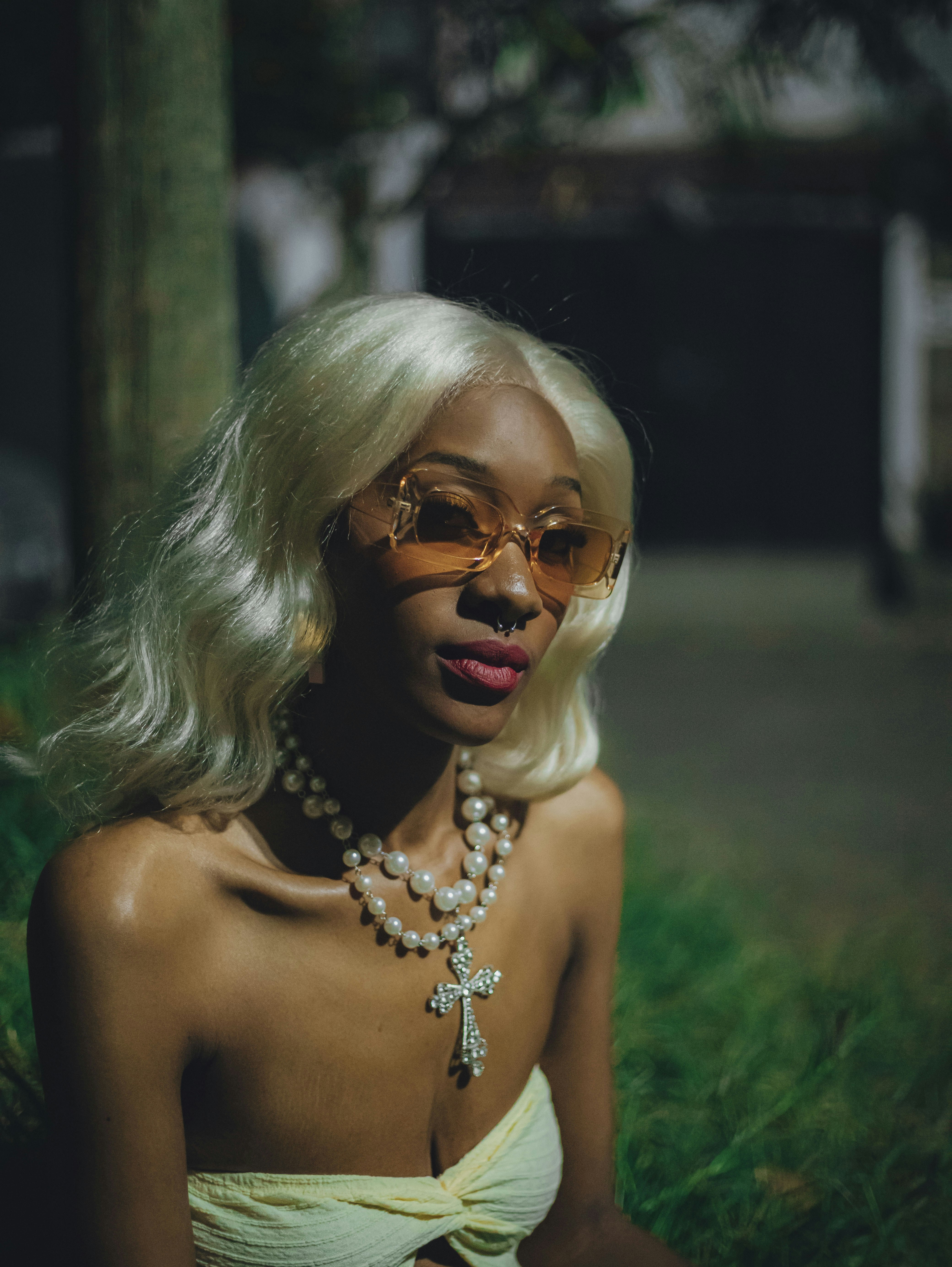 Blonde aura under shade: pearl necklace and amber lenses in a sunlit garden portrait