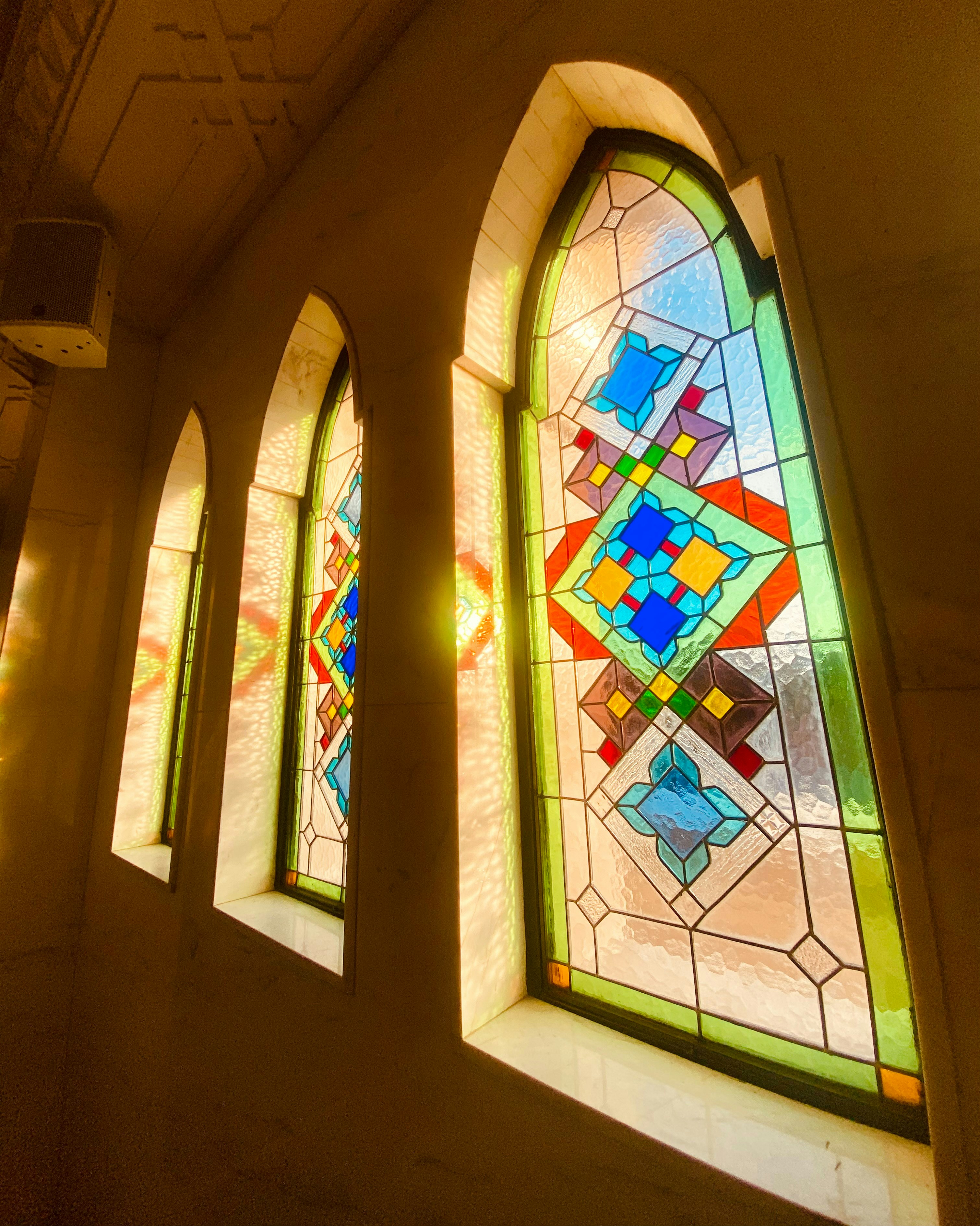 Arched stained-glass panels bathe a stone interior in warm, multicolored light, emphasizing geometric patterns and architectural depth.