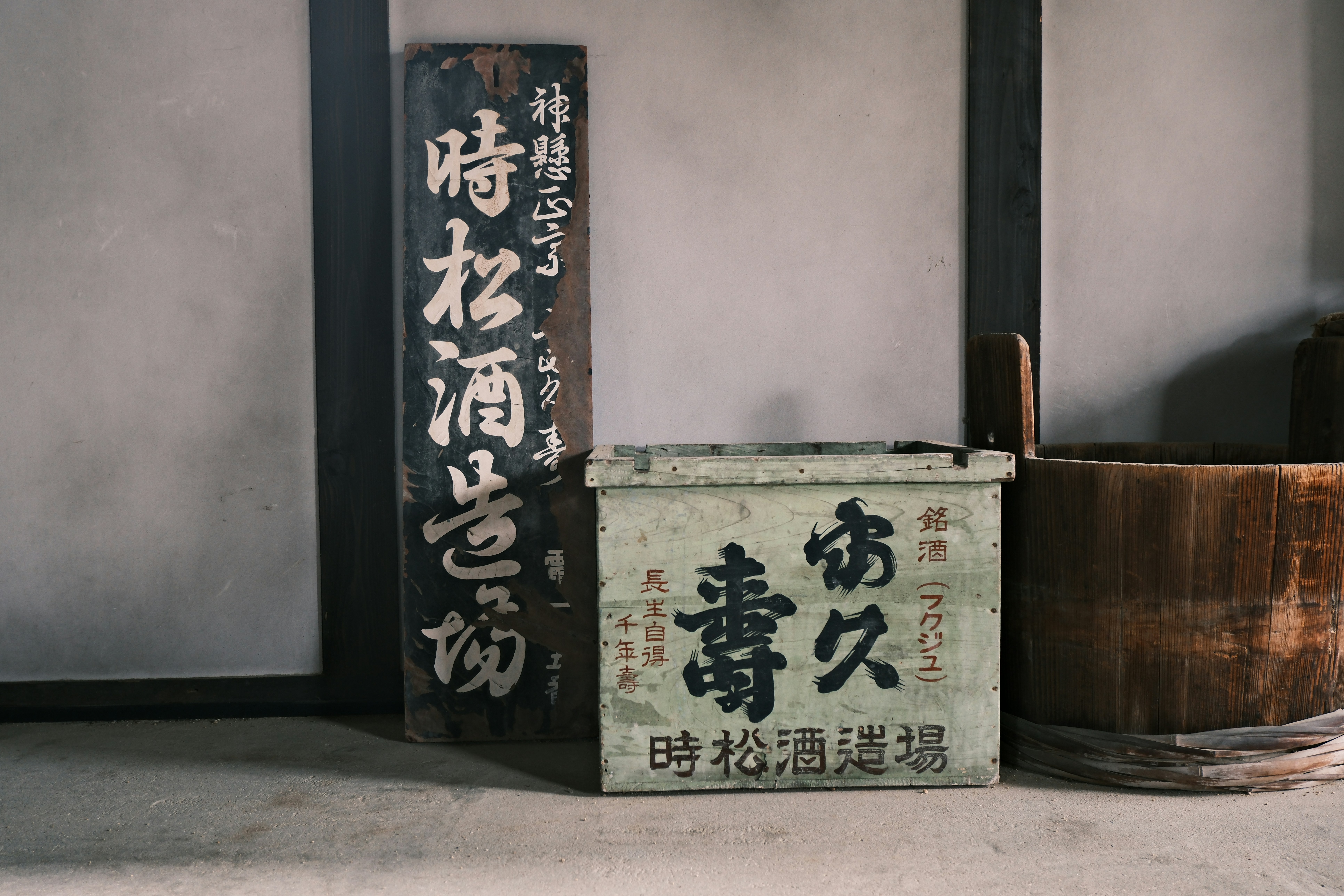 Muted storage inside a traditional space featuring two wooden crates with Japanese characters, one tall vertical plaque behind, and a curved wooden tub to the right.