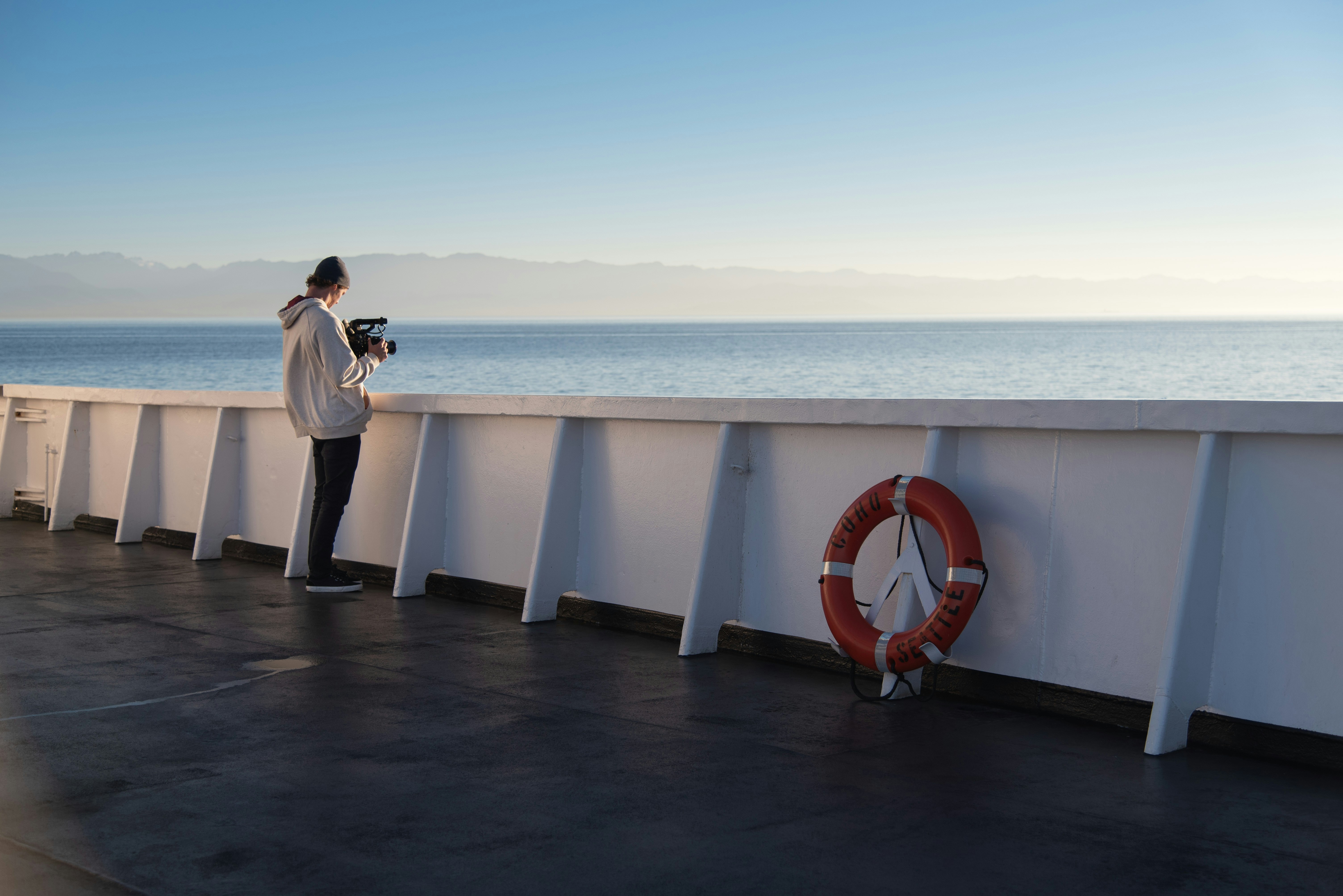 A photographer on a ship deck stands by the railing with a camera aimed toward the sea. Minimal foreground activity contrasts with the expansive ocean and distant mountains under a clear sky. The scene includes a red life ring mounted on the railing.