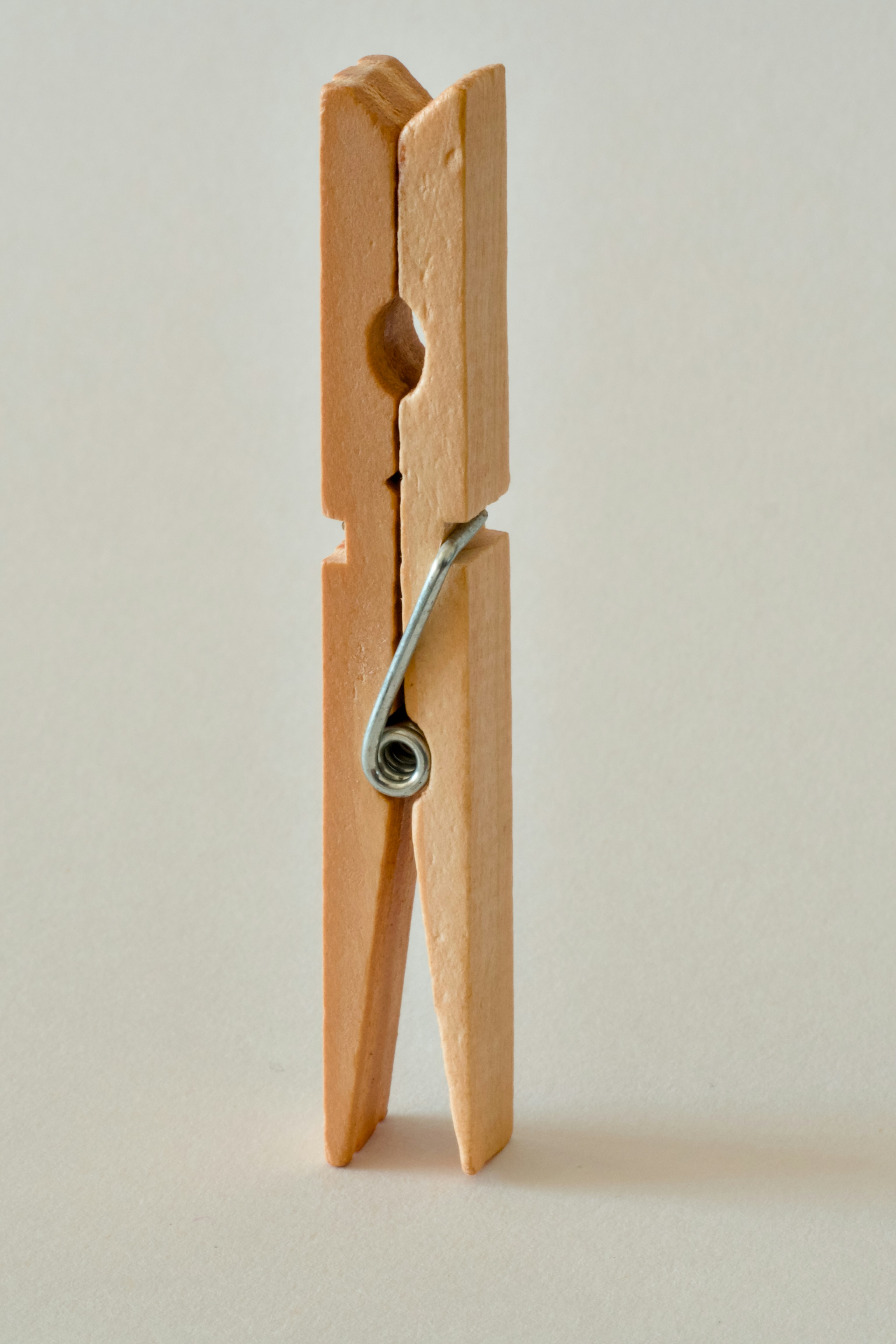 A lone wooden clothespin stands upright on a neutral surface, the metal spring loop visible along its grip. The simple still life emphasizes natural wood tones and minimalist composition.