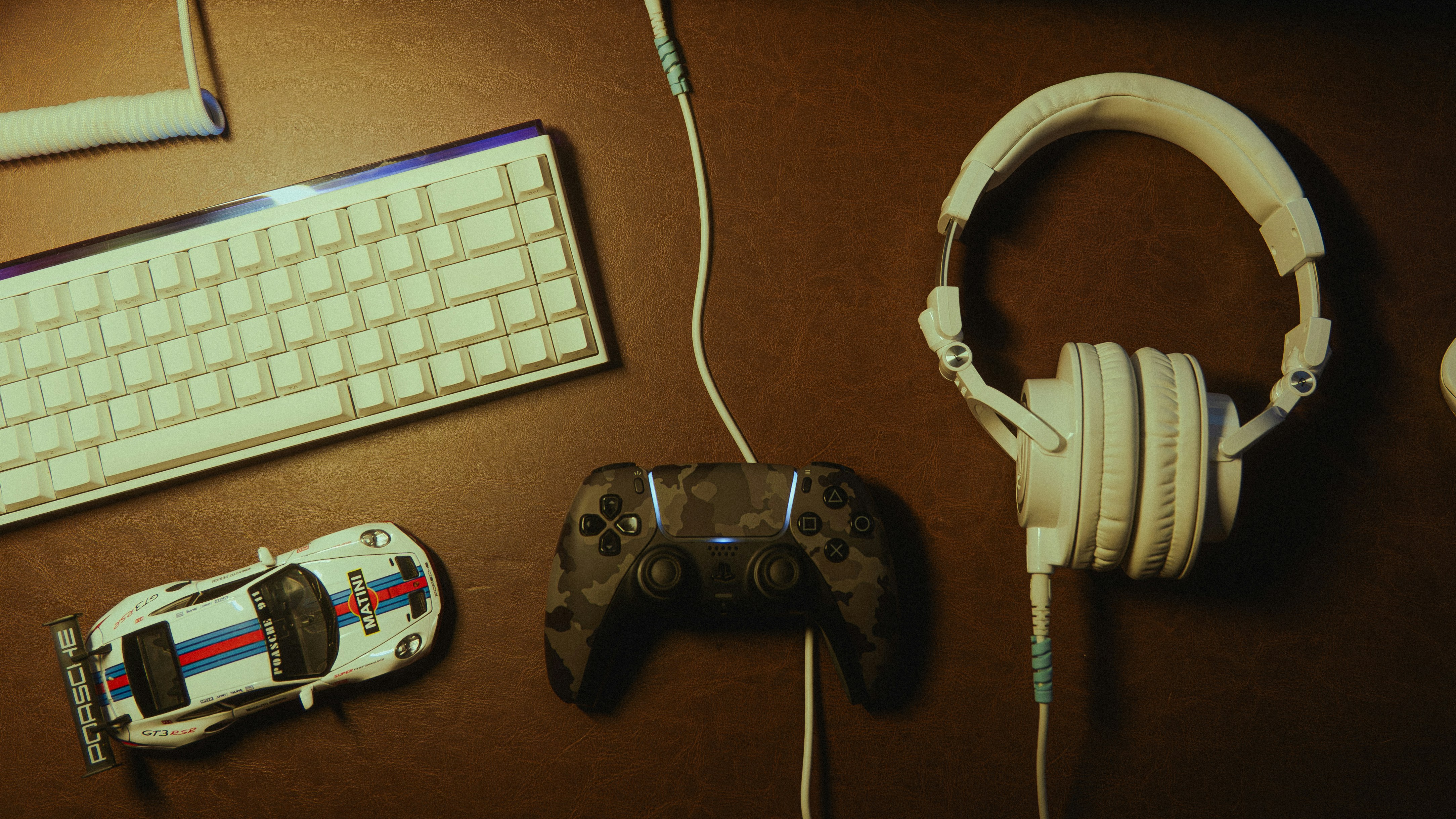 Compact desk scene with a white keyboard, a small toy race car, a black game controller, and white over-ear headphones on a brown surface.