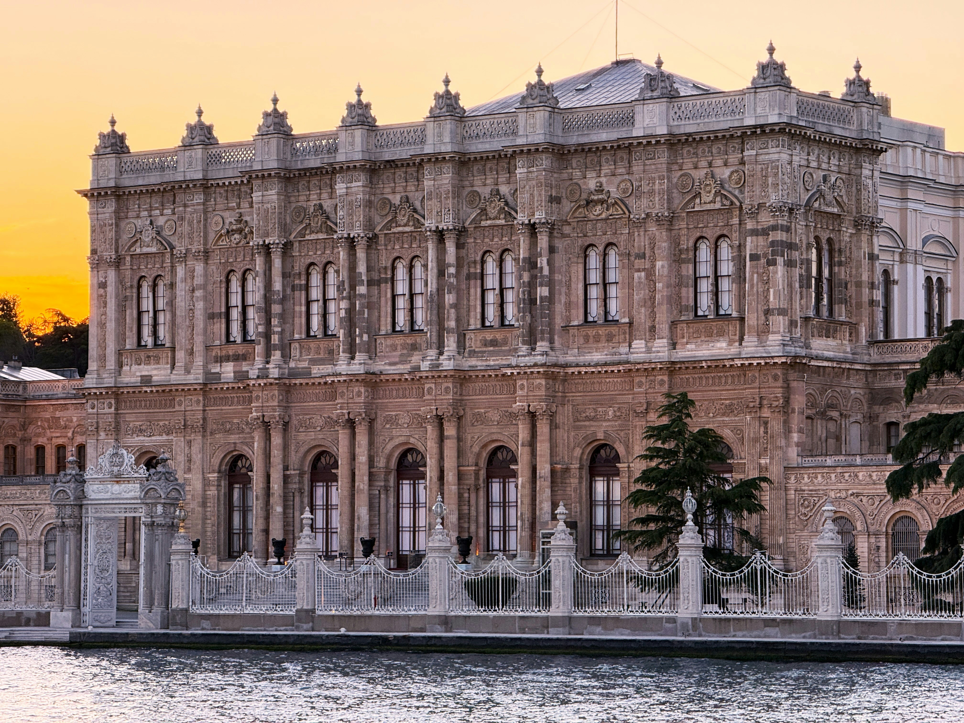 Ornate historic building along a calm waterline at sunset; warm sky highlights architectural details of the stone facade.