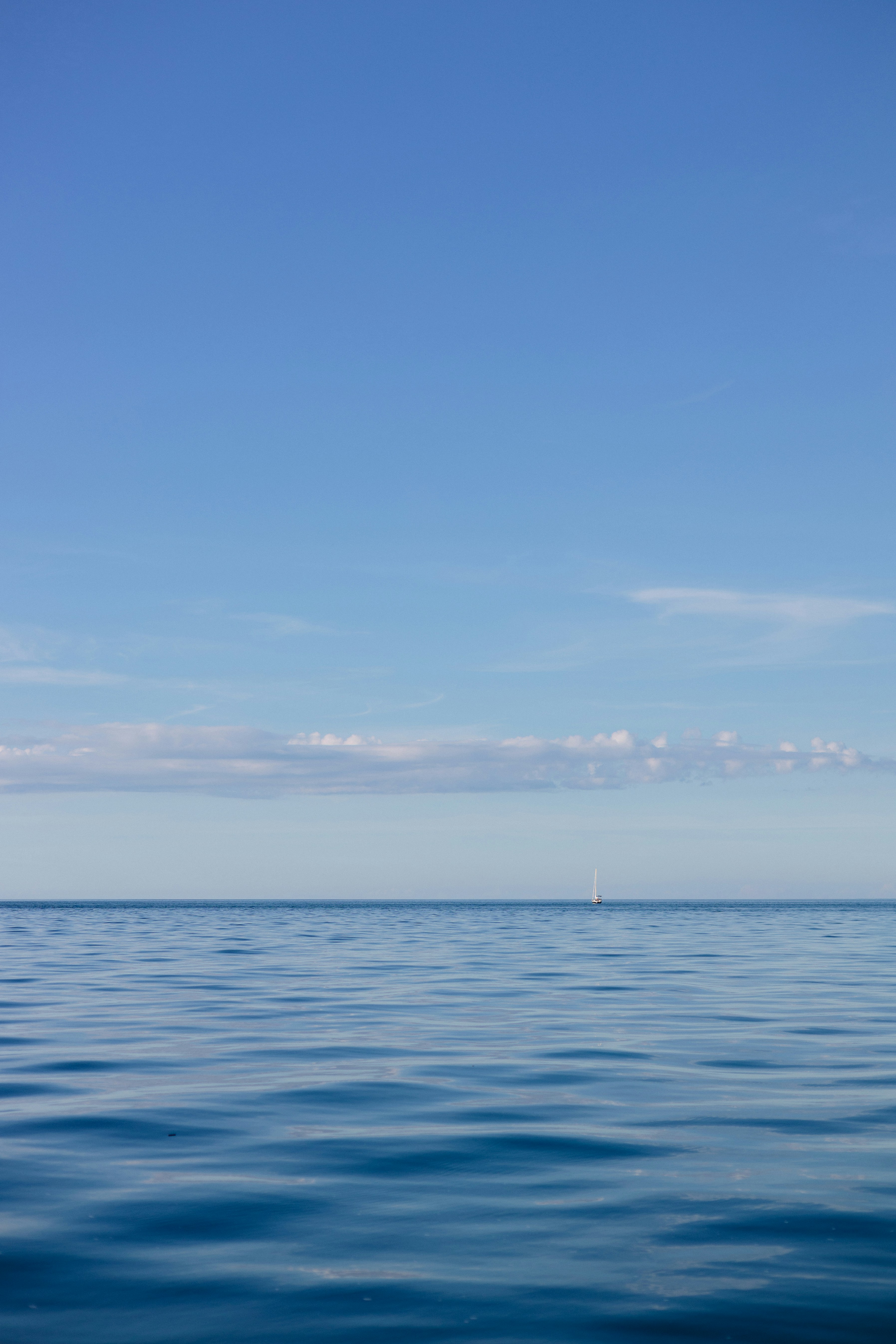 A serene seascape with a distant sailboat on a tranquil blue ocean under a clear sky. Minimalist composition emphasizing horizon line and peaceful mood.