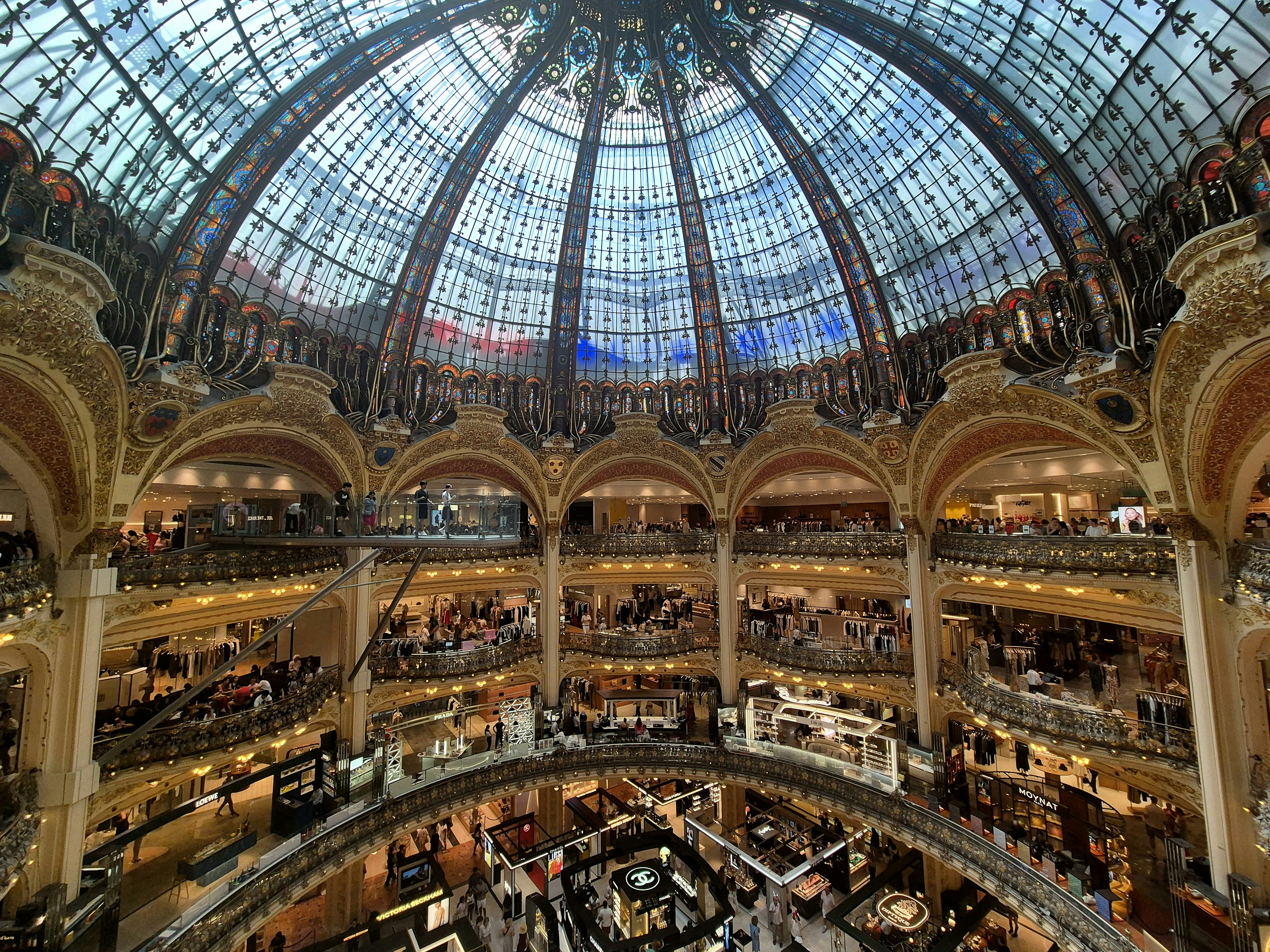 Interior of a grand multi-level shopping atrium with a glass-domed ceiling; ornate arches and balconies surround a bustling central space with shoppers and storefronts. A logo area may be present on the ground floor.