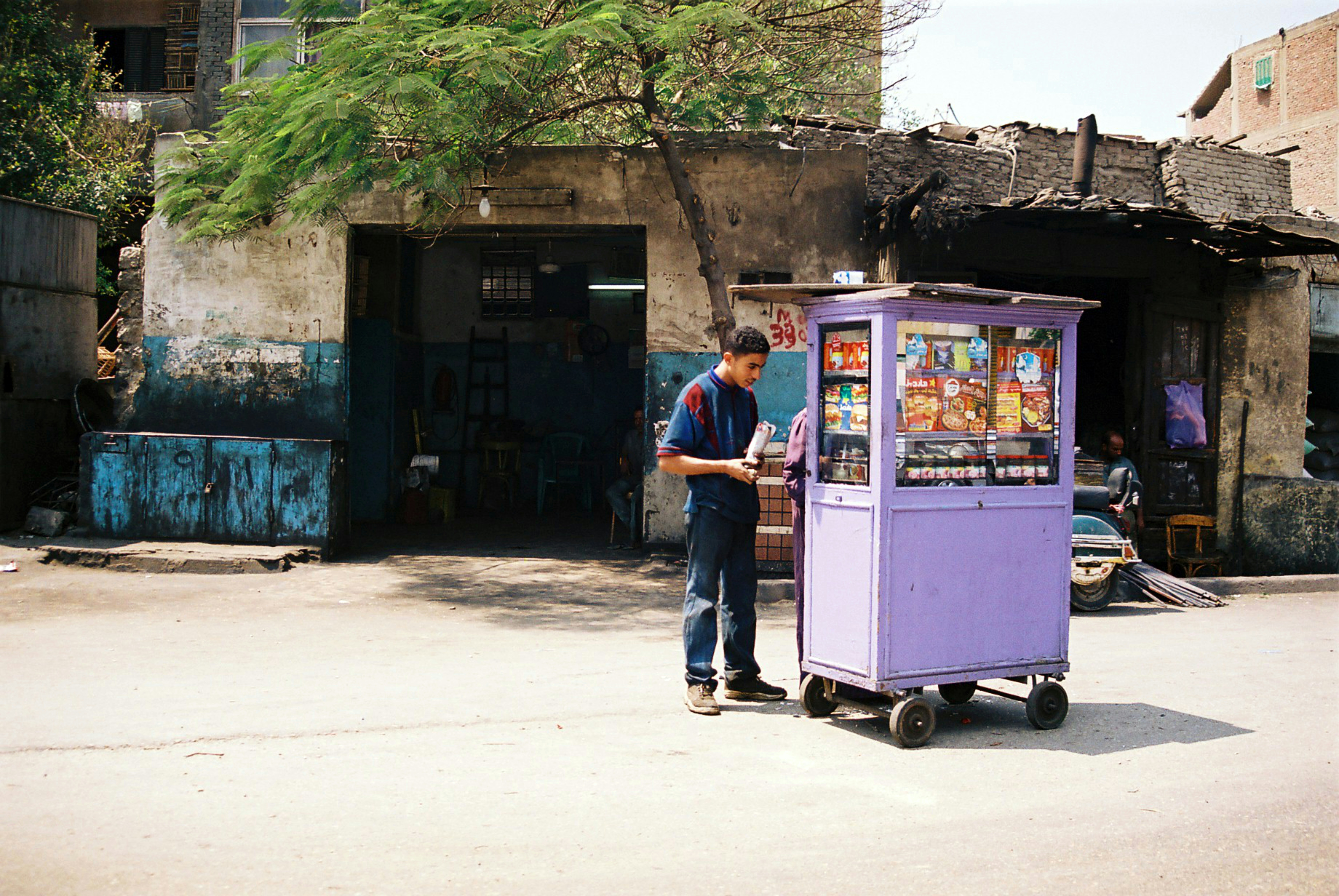 A man examines items beside a purple mobile cart against a weathered building facade; a simple street scene highlighting a food vendor setup.