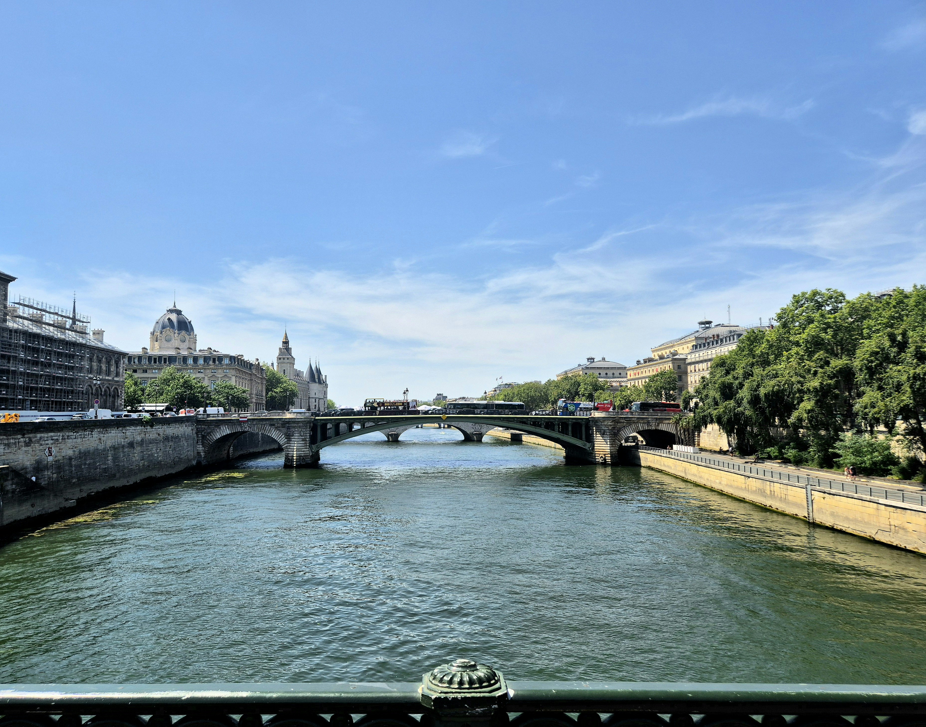 Wide river scene along a historic city canal with stone bridges and riverside façades. A foreground railing frames calm water and arches under the bridges, capturing urban architecture and blue sky.
