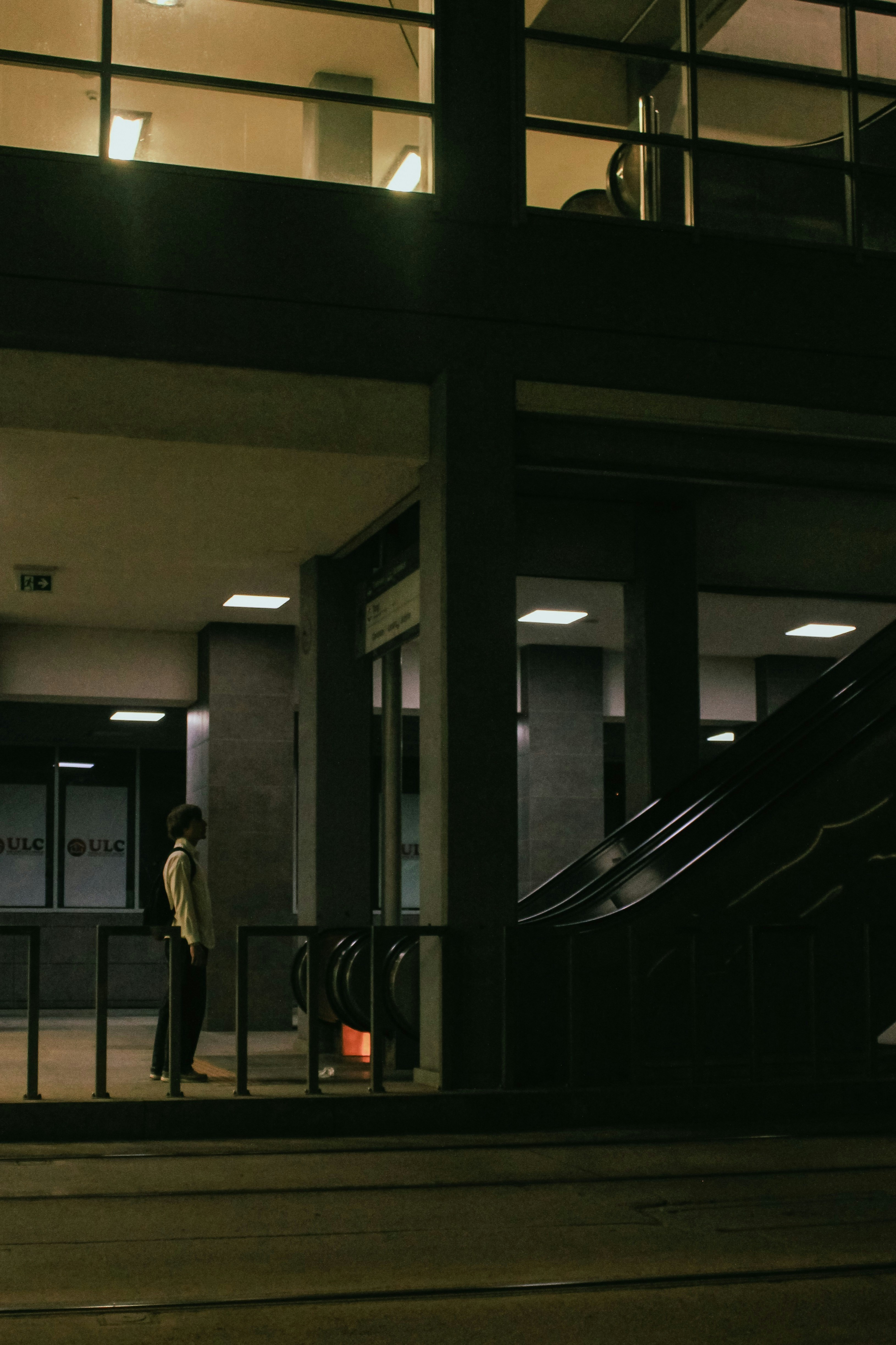 Figure in a Quiet Plaza: A Person Waiting by the Escalator Under Dim Urban Lighting
