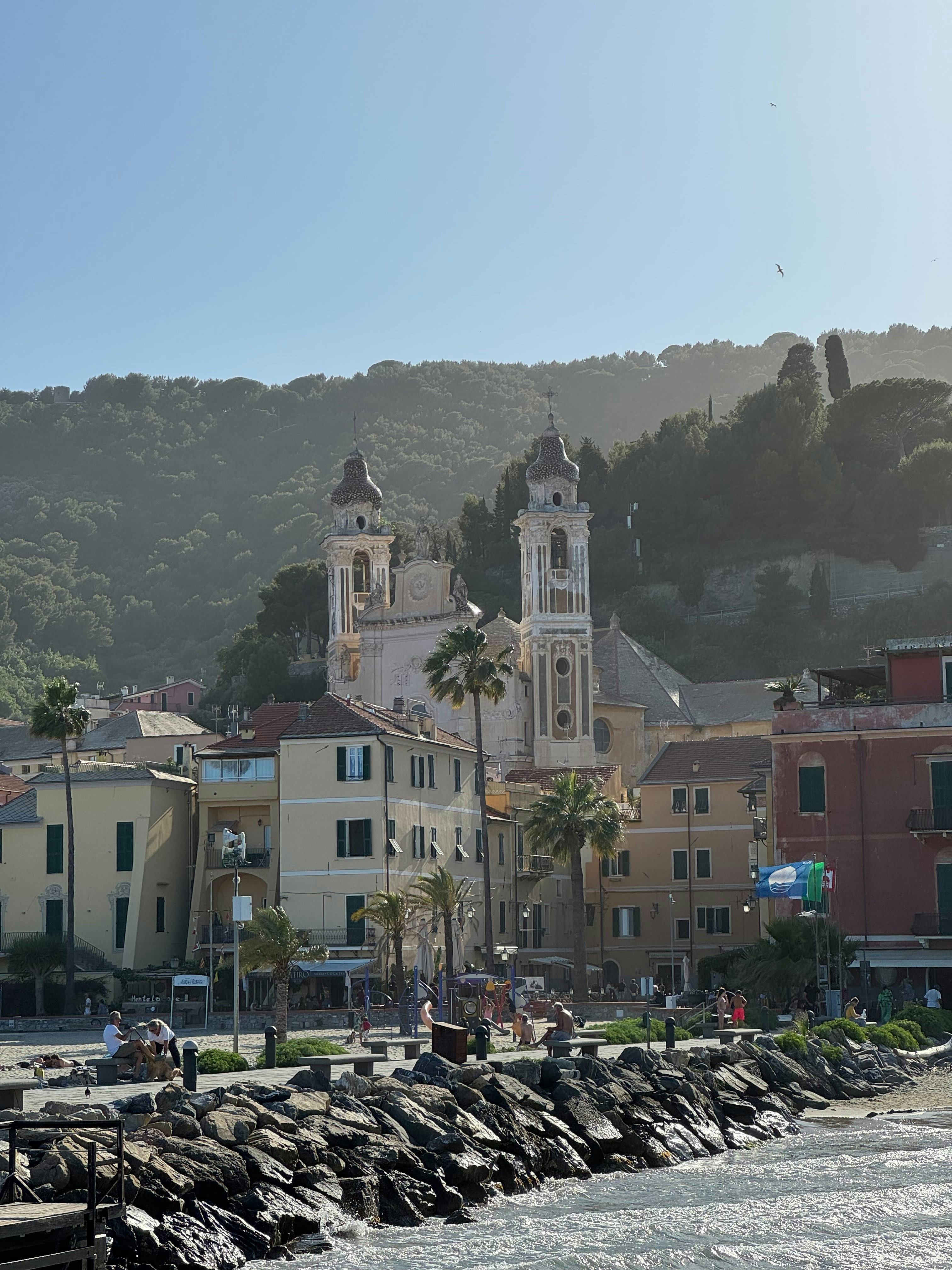 Sunlit coastal townscape with a prominent twin-towered church behind a row of pastel buildings along the rocky waterfront.