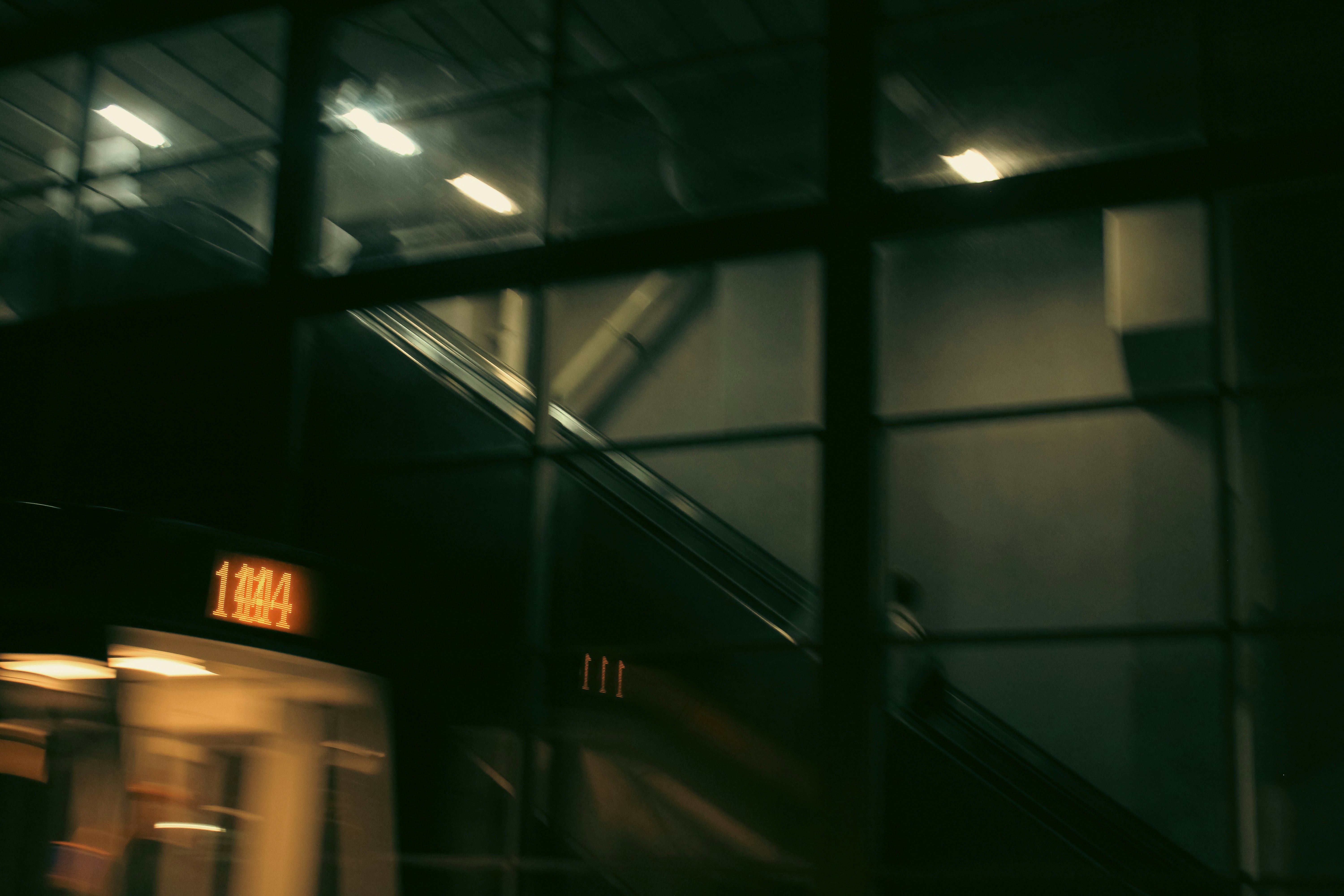 Blurred indoor scene with an escalator and a glowing train indicator reflected through a glass facade; abstract urban motion with shallow depth and warm tones.