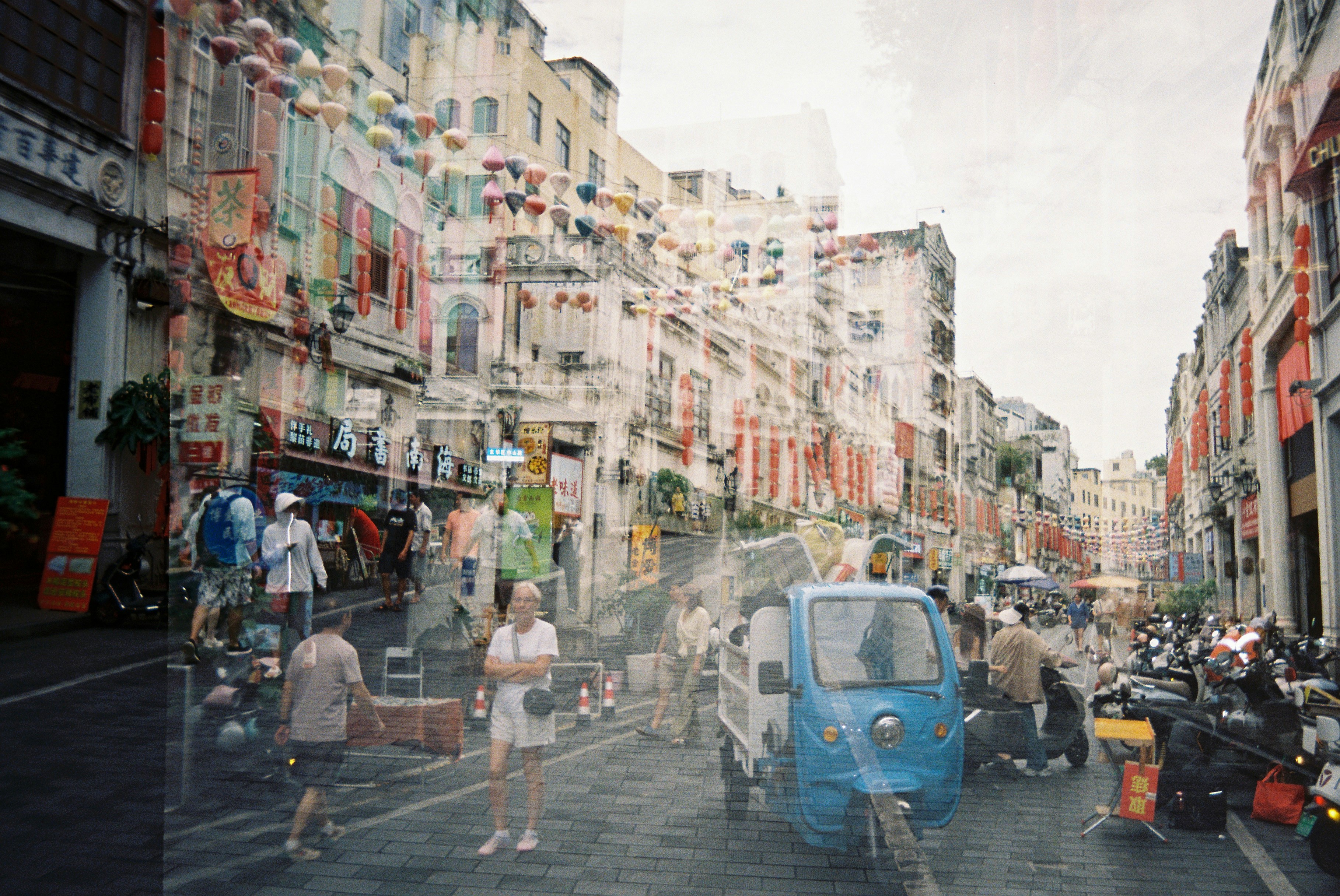 A double-exposure film photograph of an old colonial-era street in a city on Hainan Island, China. The overlay of two nearly identical frames creates a ghosting, layered effect typical of analog photography. The colonial architecture, arcades, and nostalgic atmosphere are intensified by the dreamy and surreal quality of the unintended double exposure.