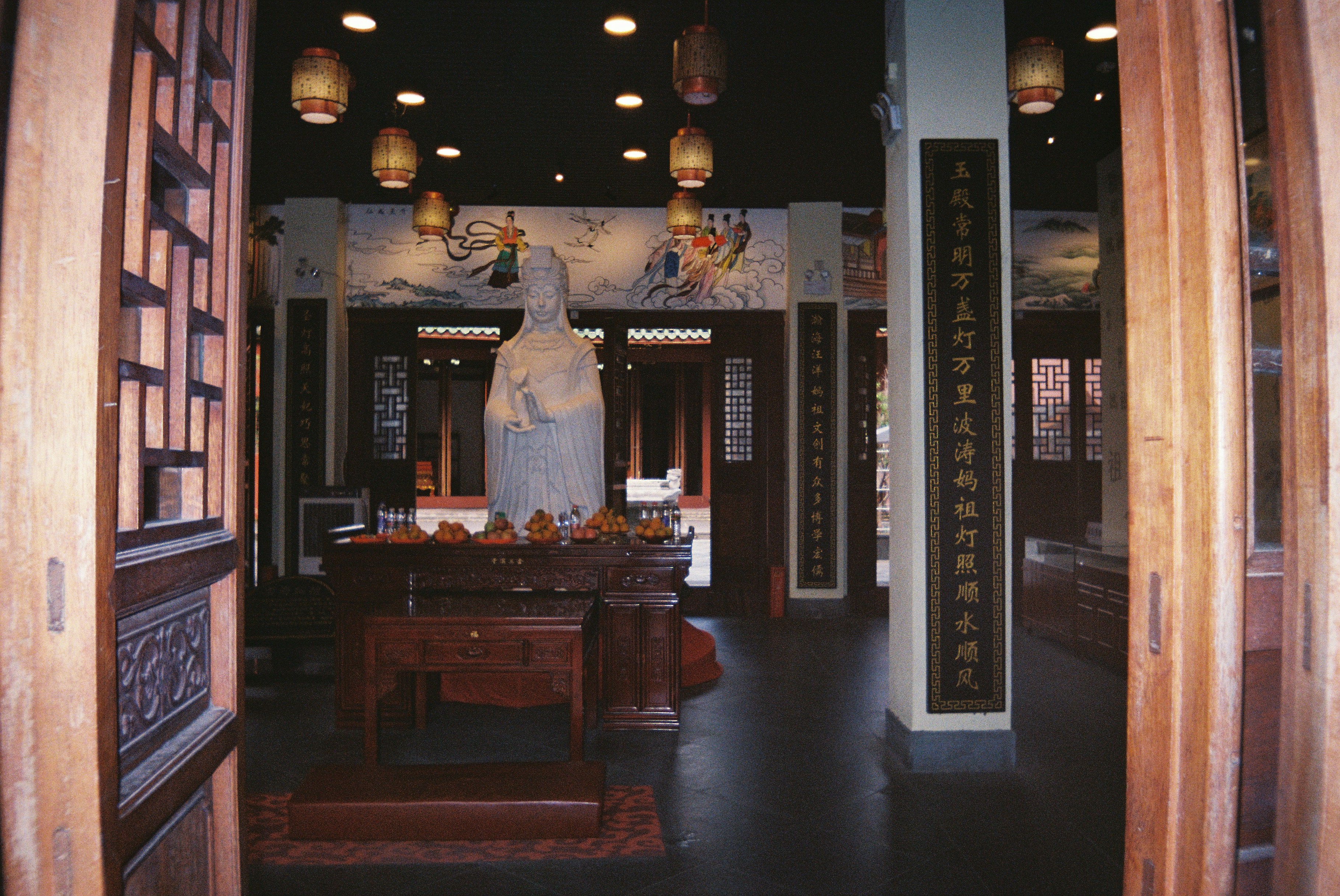 The entrance to a Buddhist temple on Hainan Island, dedicated to a revered local goddess. Inside, a statue of the deity, possibly Guanyin, Mazu, or Shui Wei Sheng Niang, is visible along with offerings placed nearby. The scene captures the spiritual atmosphere and cultural richness of this sacred island shrine.