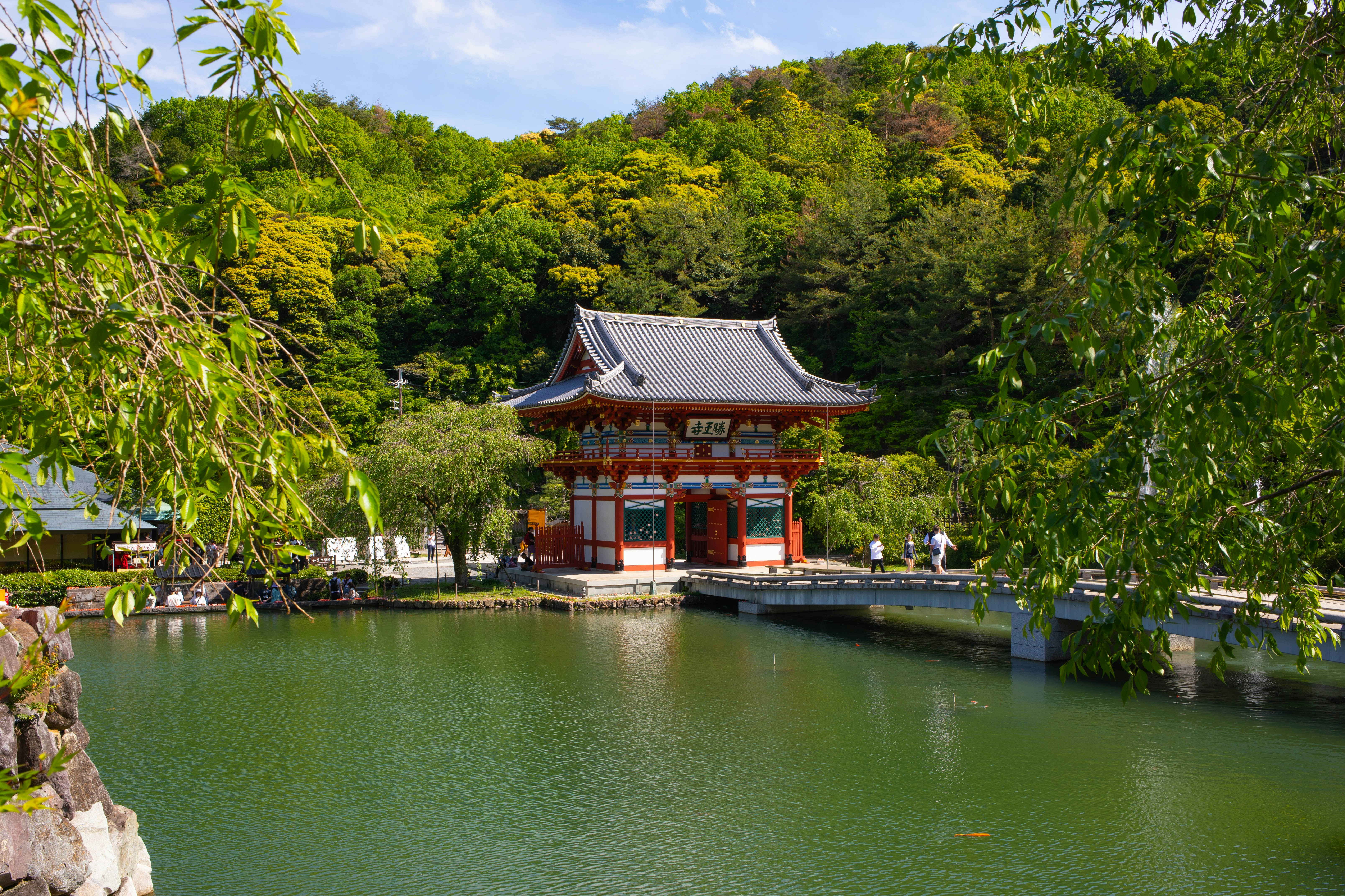 Stone-and-wood temple sits on a calm pond, framed by lush trees and a distant hillside, inviting a serene moment of contemplation.