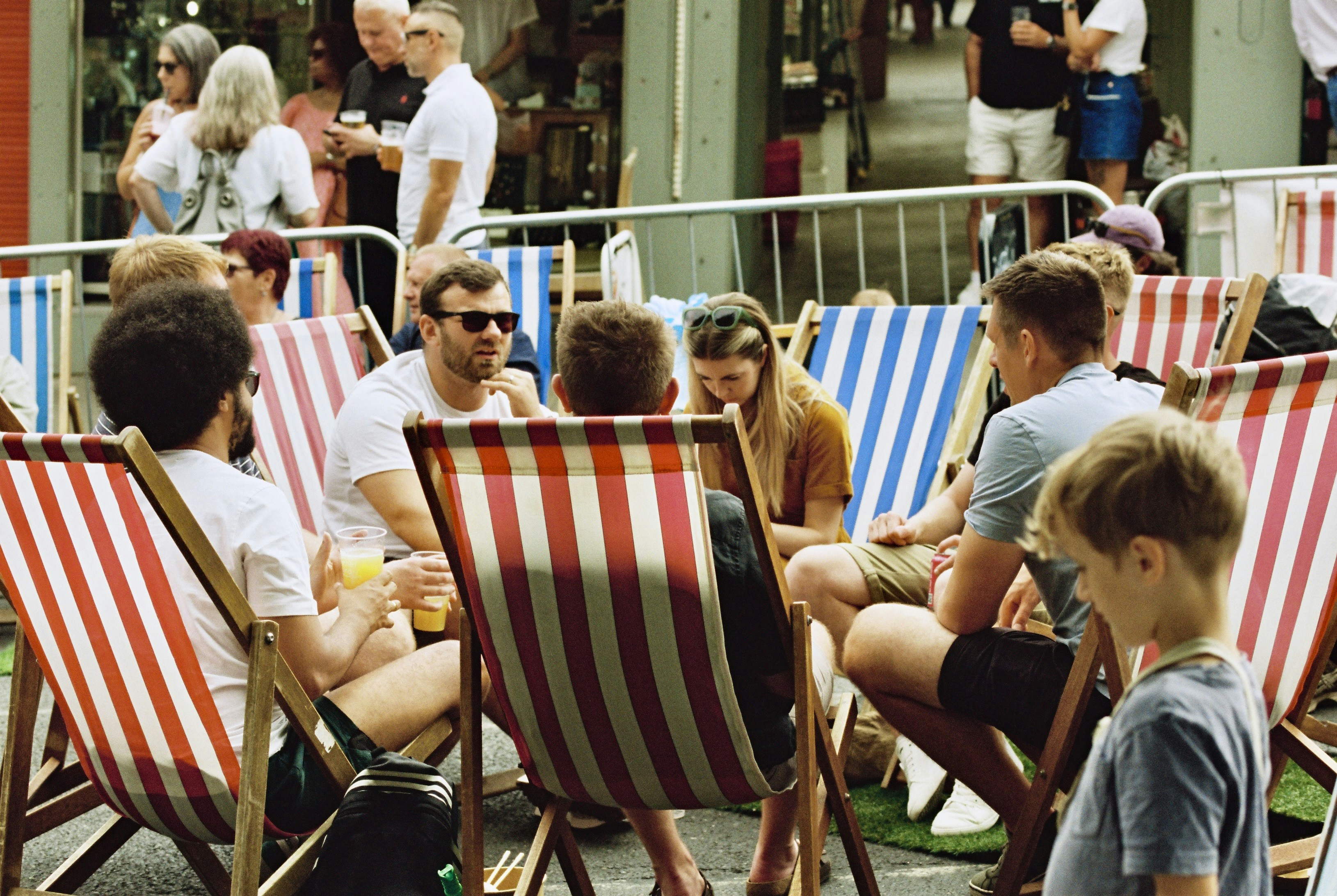 Friends relax with cold drinks in red-and-white striped deckchairs at a busy summer street event.
