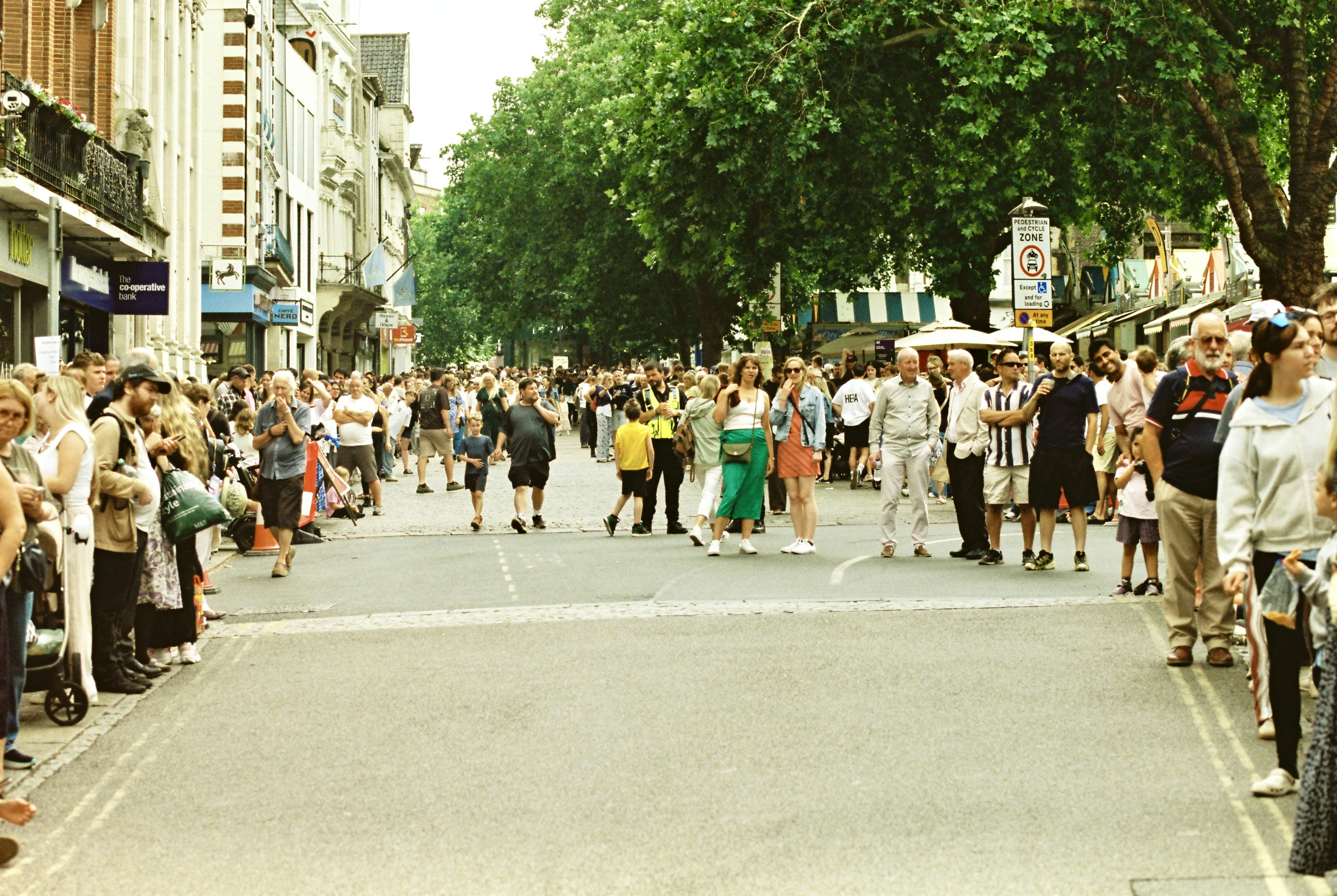 Crowds fill a pedestrianised city high street lined with shops and leafy trees on a summer festival day.