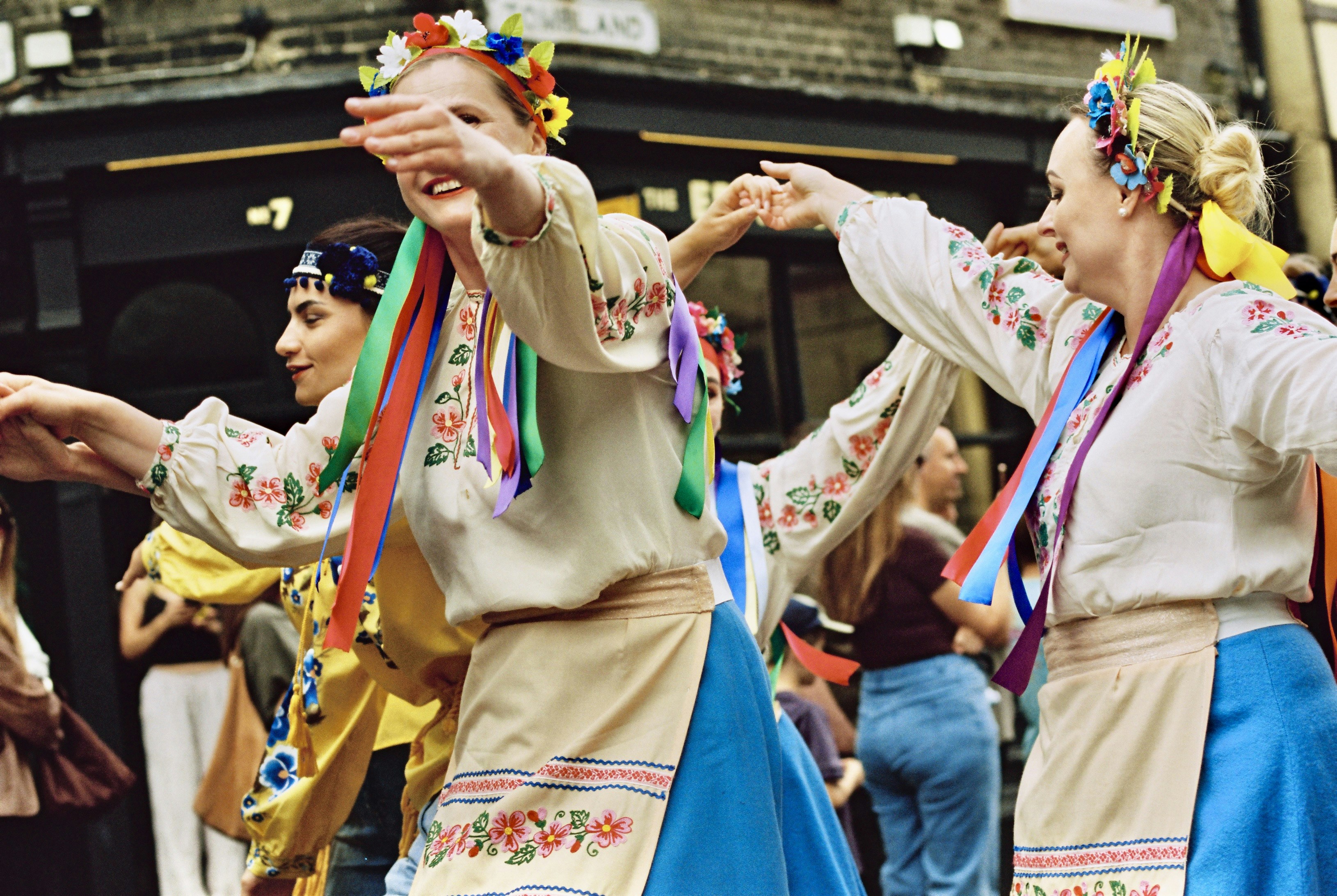 Dancers in embroidered folk costumes and flower crowns swirl coloured ribbons through a street festival.
