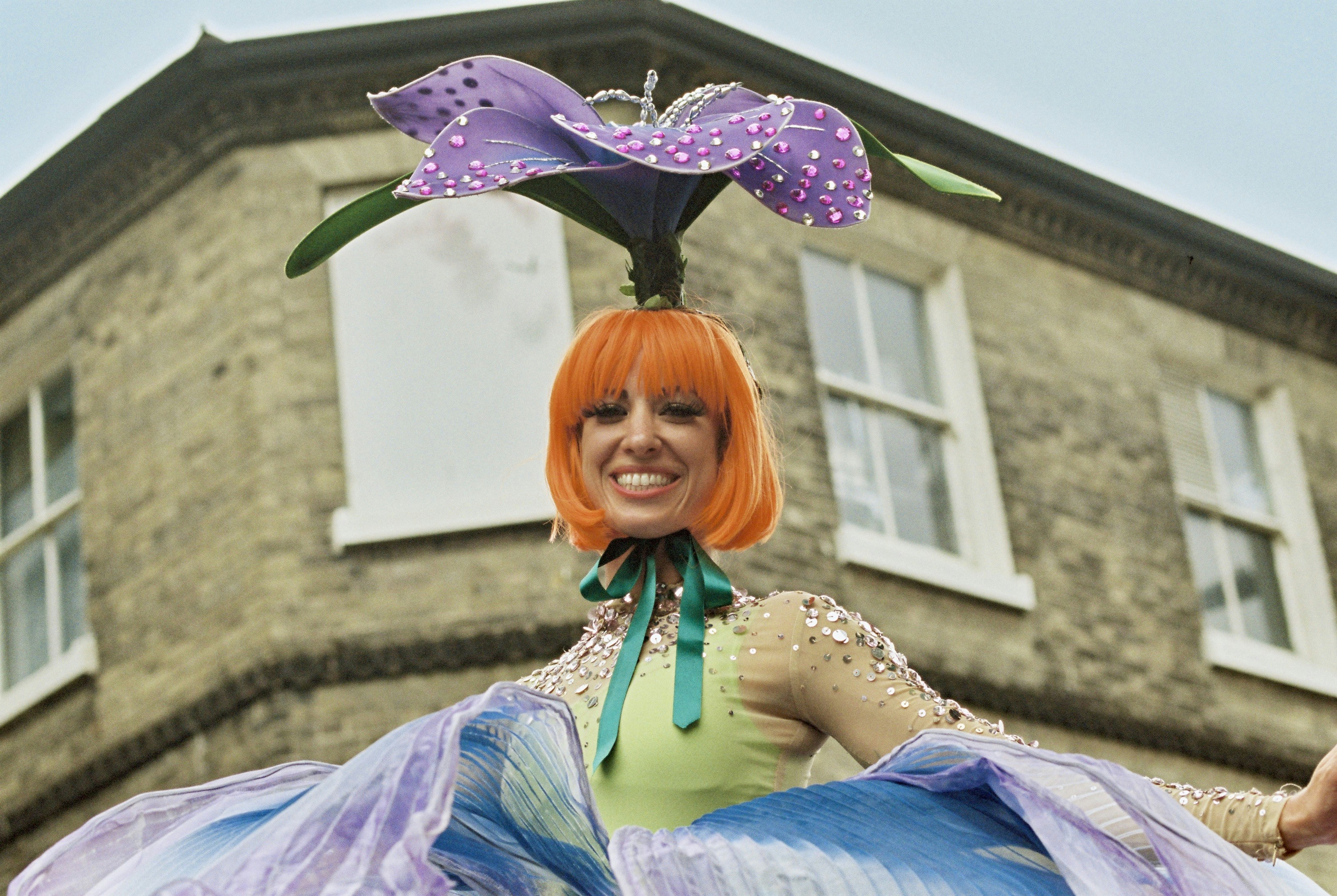 A stilt walker in a jewelled bodysuit and purple flower headdress smiles above the summer carnival crowd.